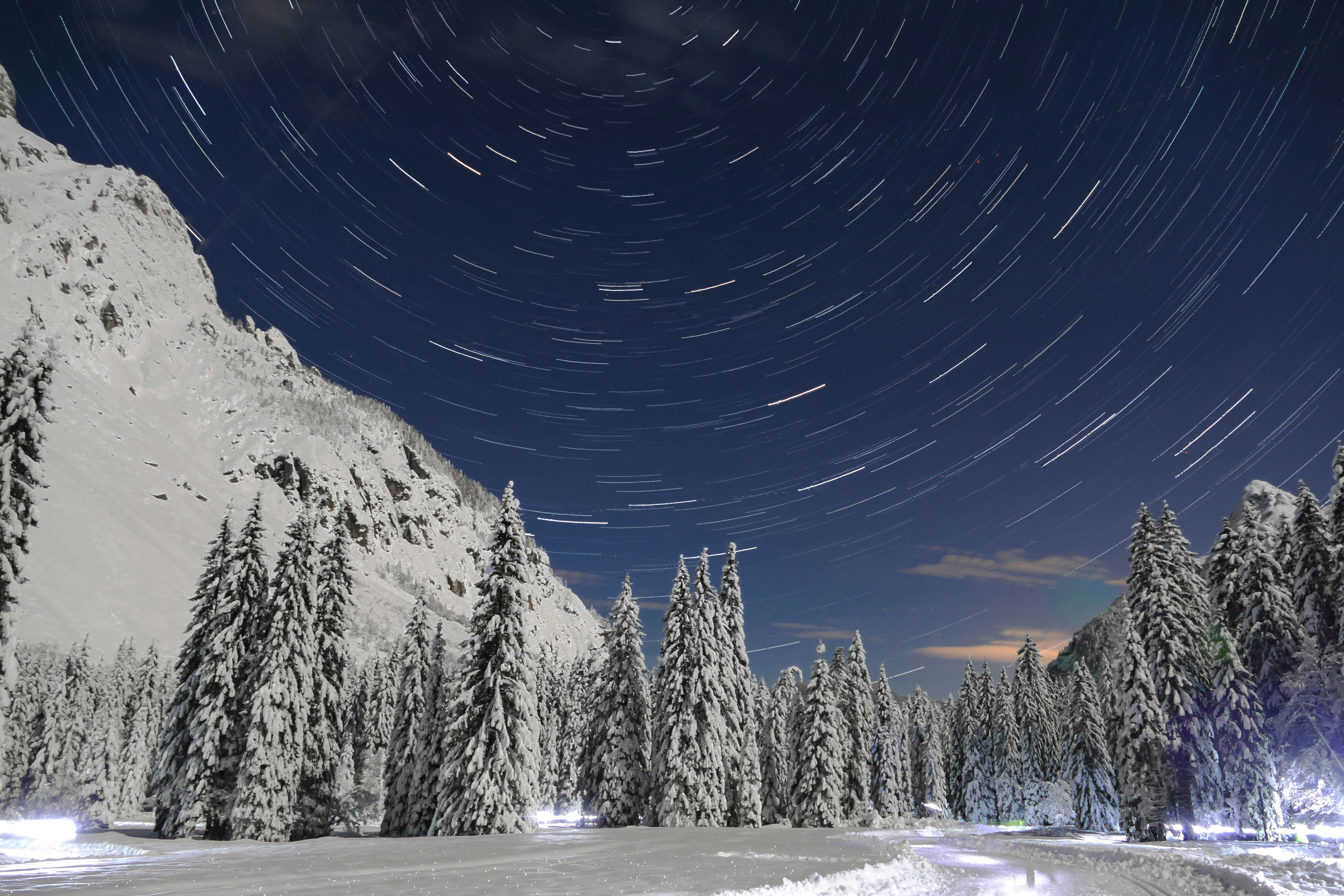 Star trails swirl over a snow-covered forest, illuminated by a moonlit sky and distant lights. The scene captures the serene beauty of winter nights in nature.