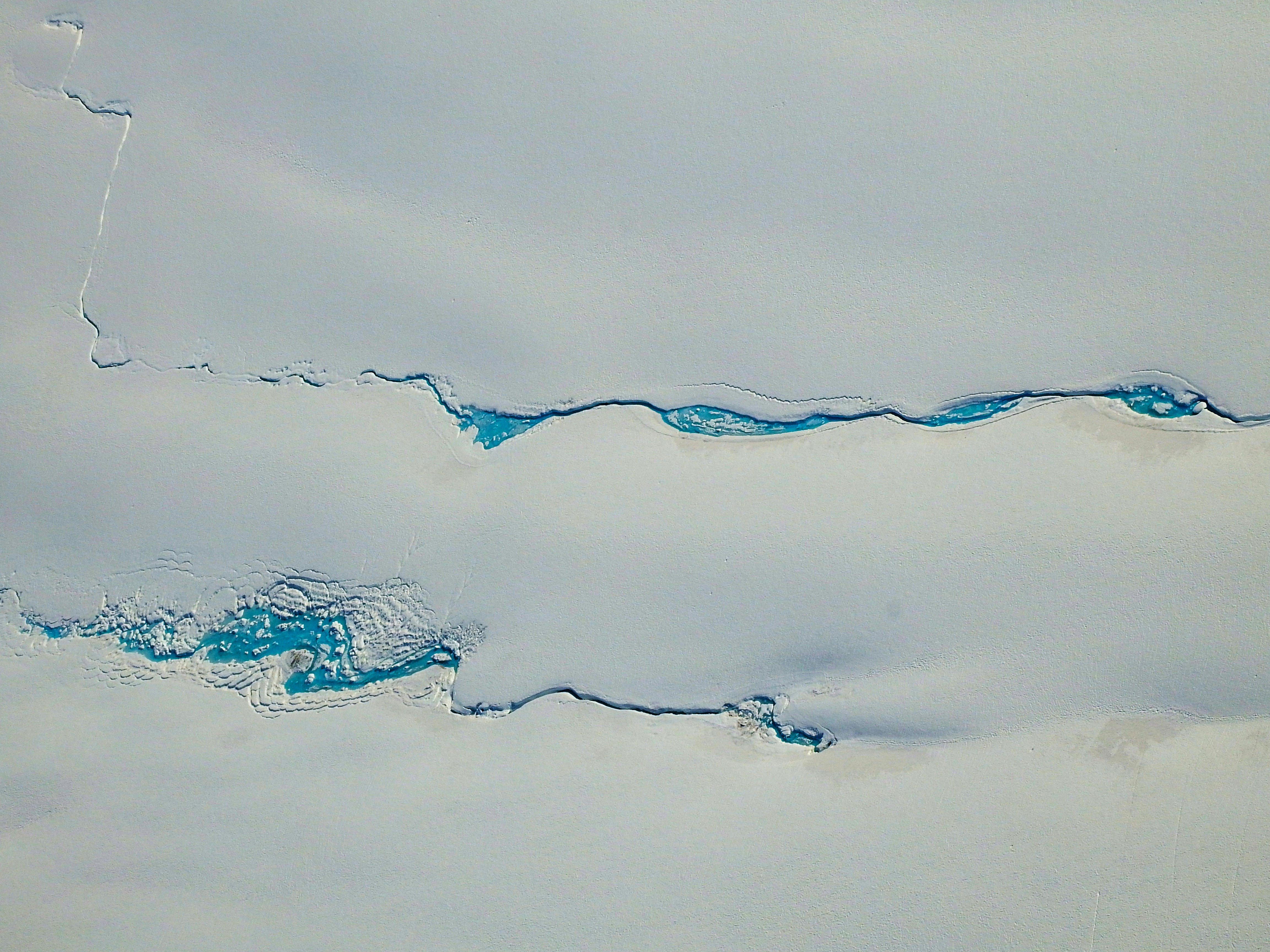 Aerial view of a glacial landscape revealing cracks filled with vibrant blue water amidst vast white ice. The stark contrast highlights the dynamic nature of the frozen environment.
