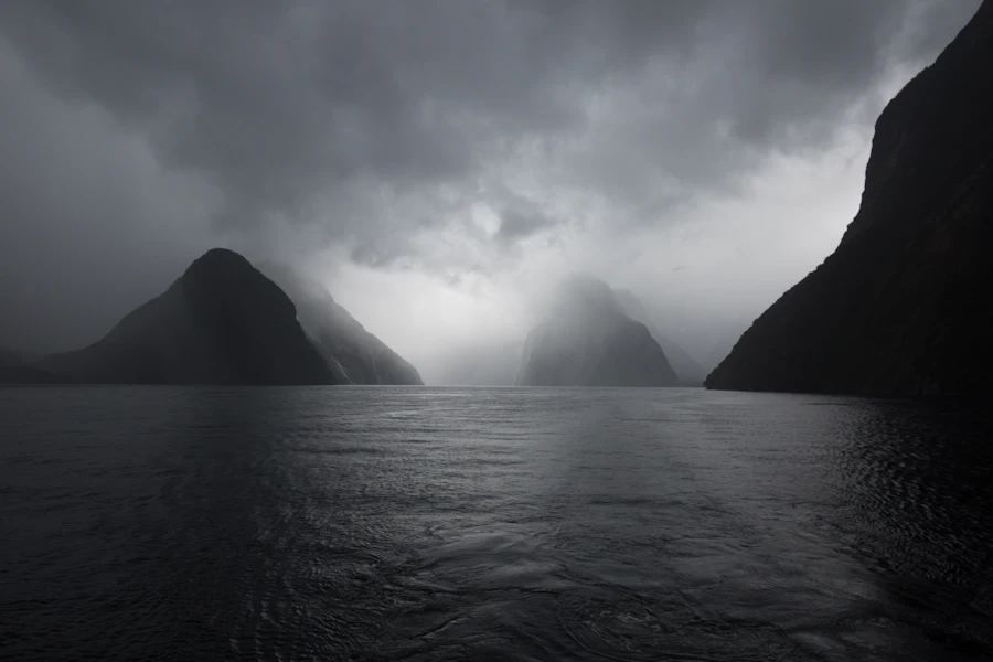 A moody seascape with steep black mountains framing a narrow channel of water under heavy storm clouds. Bright mist glows at the horizon between the peaks.