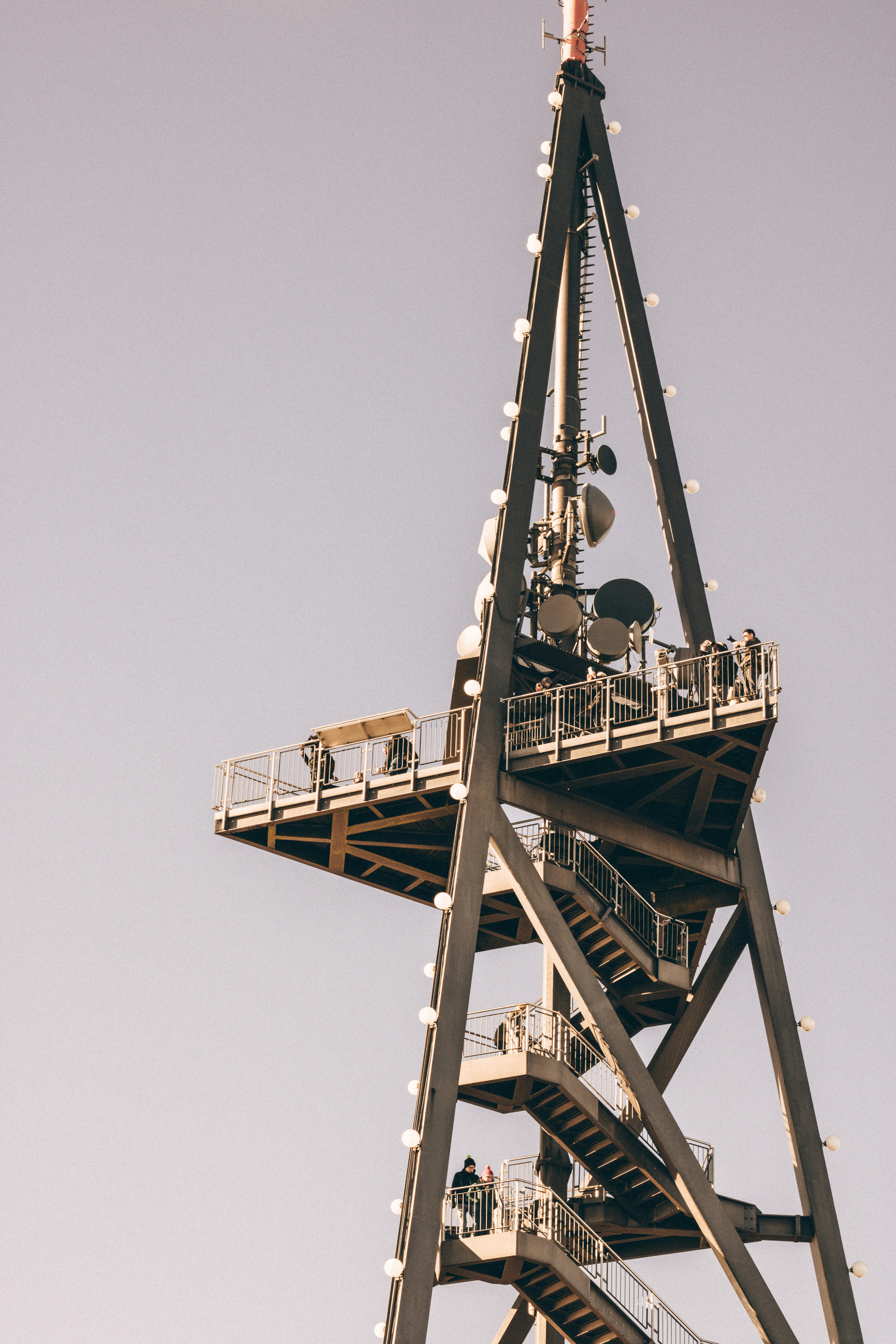 Telecommunications tower with a spiral staircase and observation deck, showcasing individuals enjoying the view. Lights adorn the structure against a pastel sky.