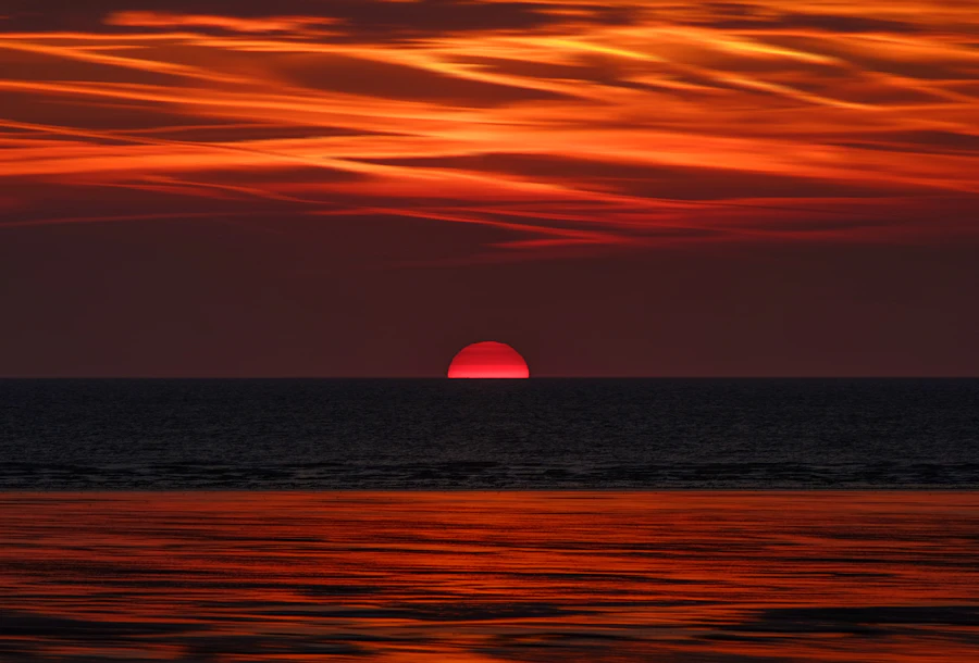 Half sun rising above calm ocean with vibrant red clouds overhead.