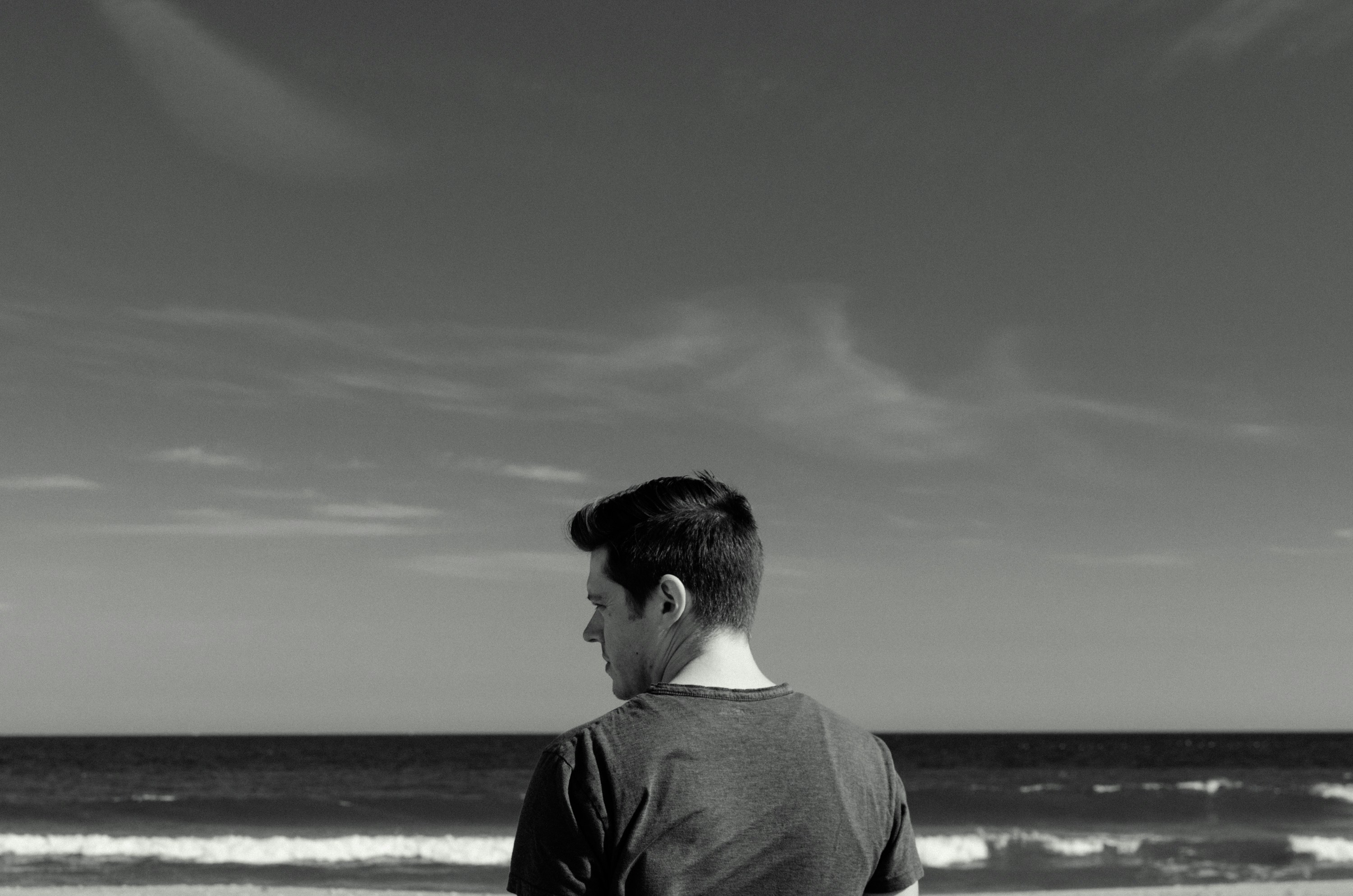 Person standing on a beach looking out at the ocean under a cloudy sky.