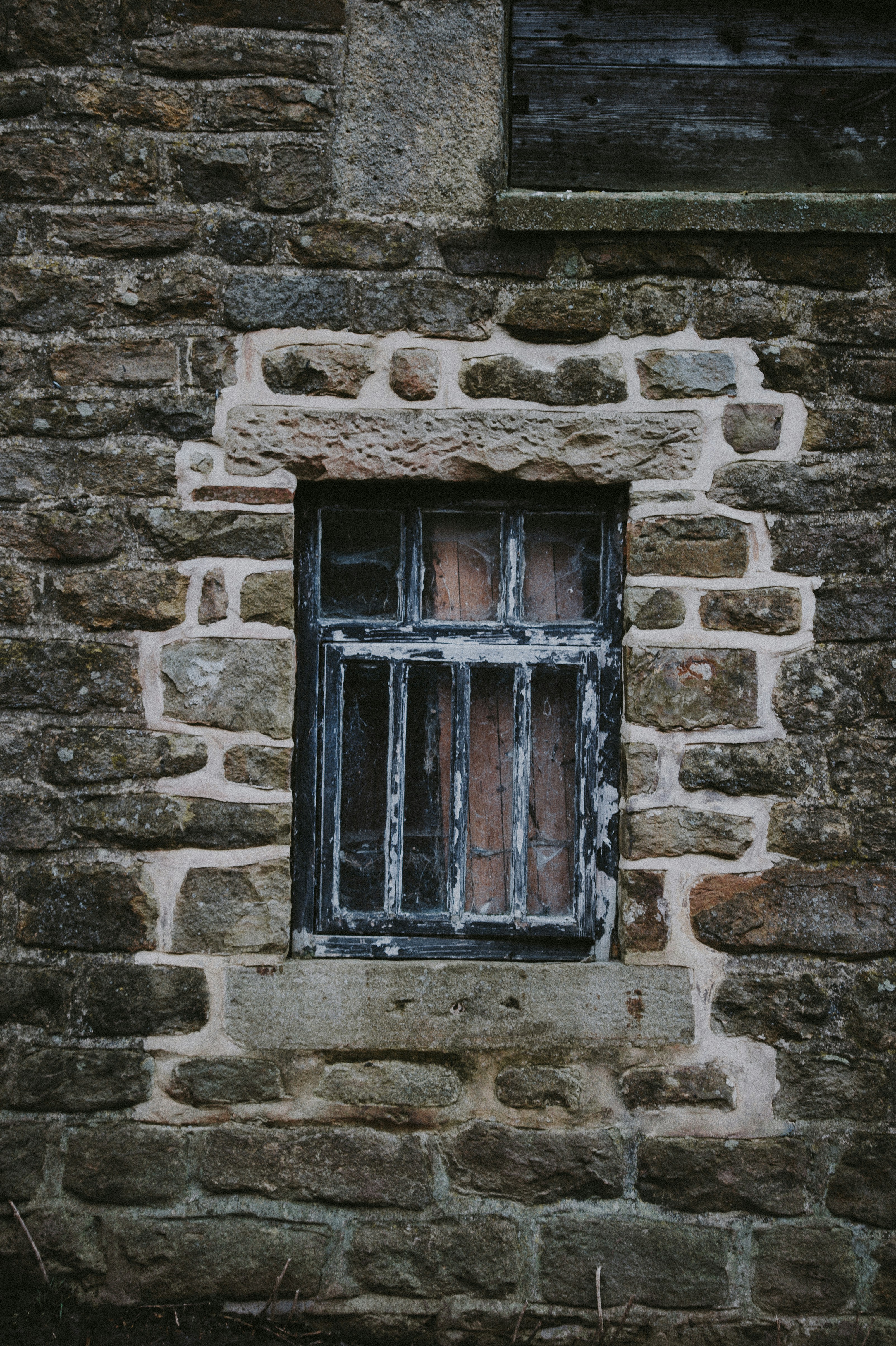 A weathered window framed by rough stone walls, showcasing the passage of time and nature's embrace. The peeling paint and muted colors evoke a sense of nostalgia.
