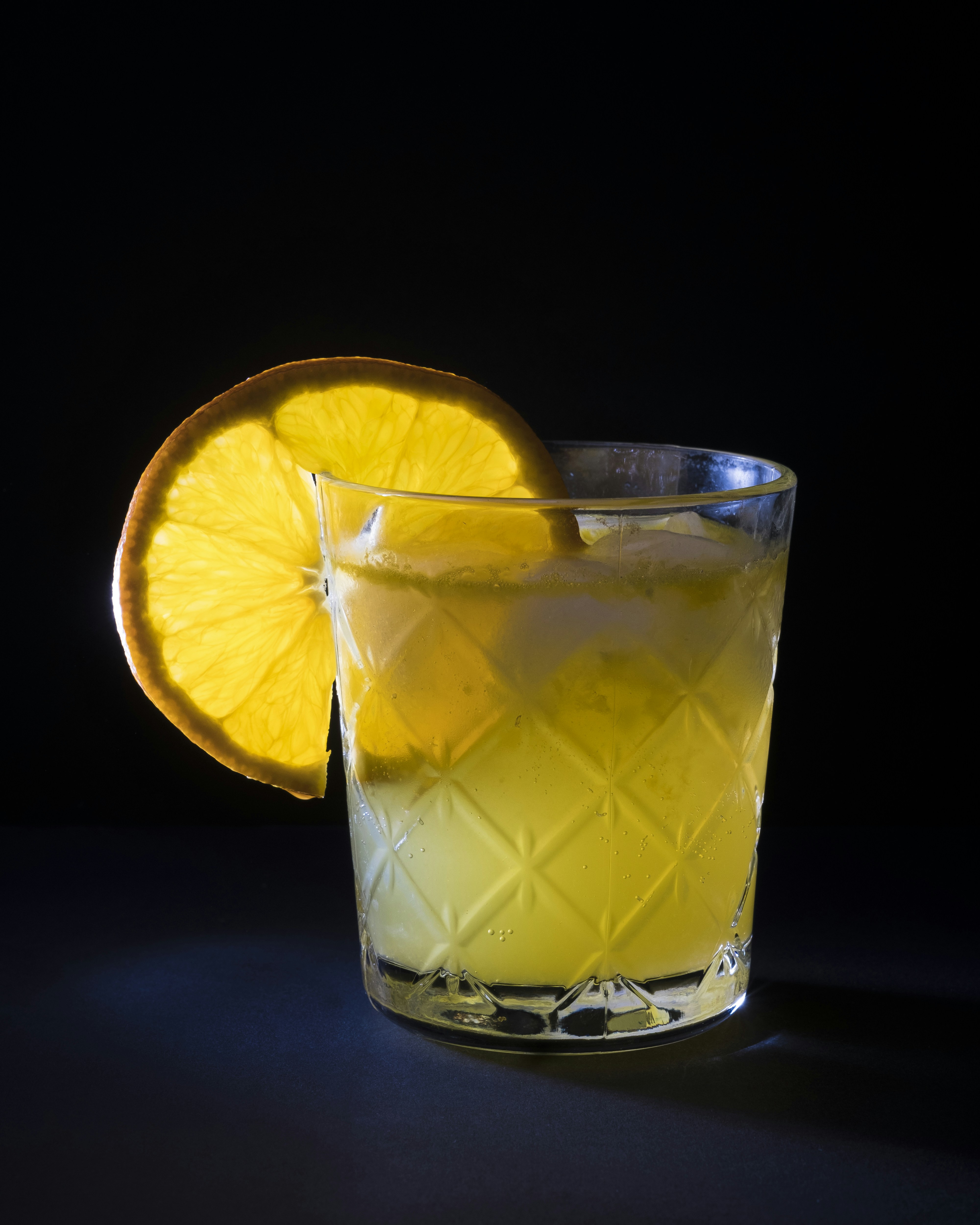 A vibrant yellow drink garnished with a fresh orange slice, set against a dark backdrop, highlighting its refreshing appeal.