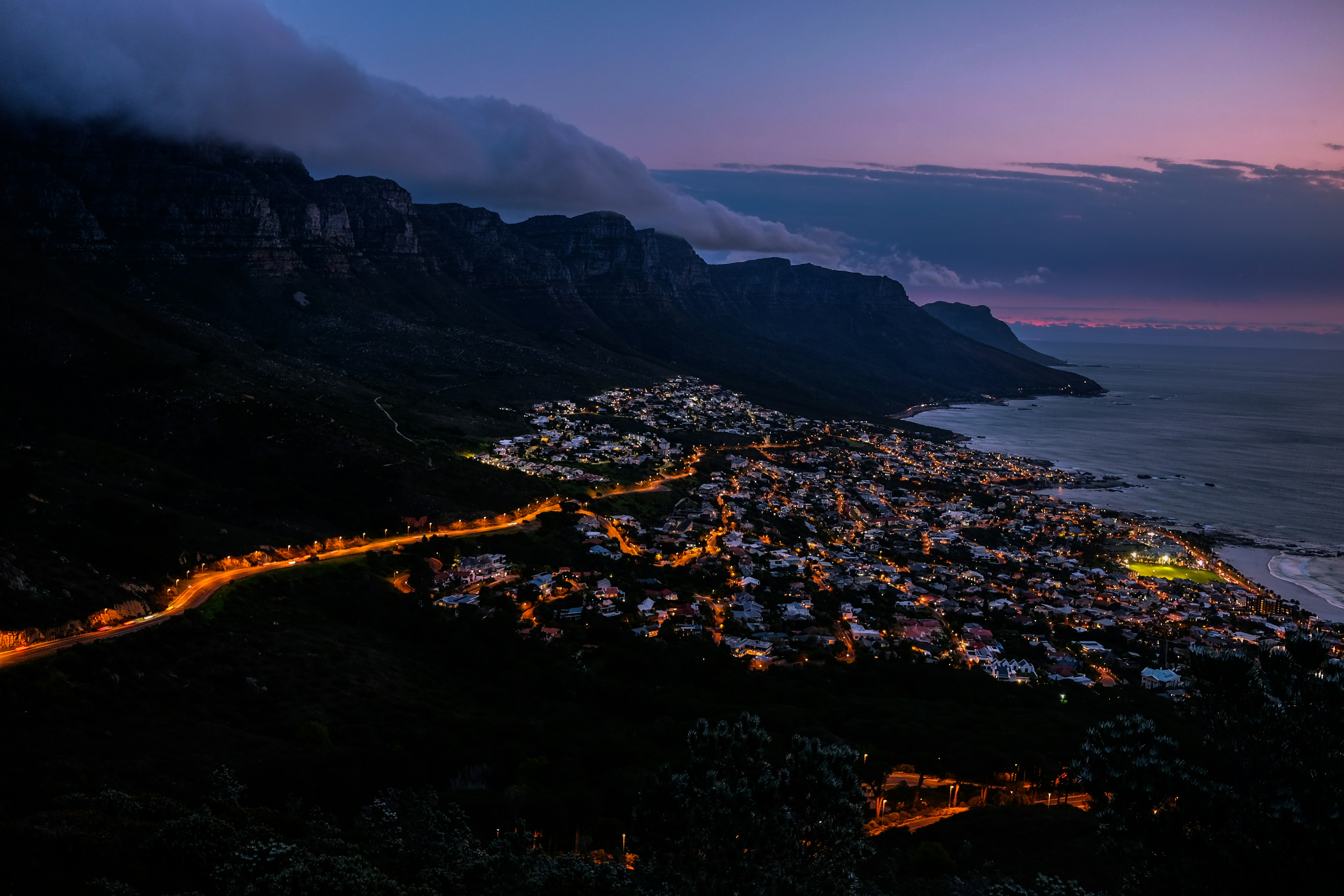 Aerial photograph of a coastal town at blue hour, with a serpentine road of orange streetlights winding through foothills toward the shoreline.