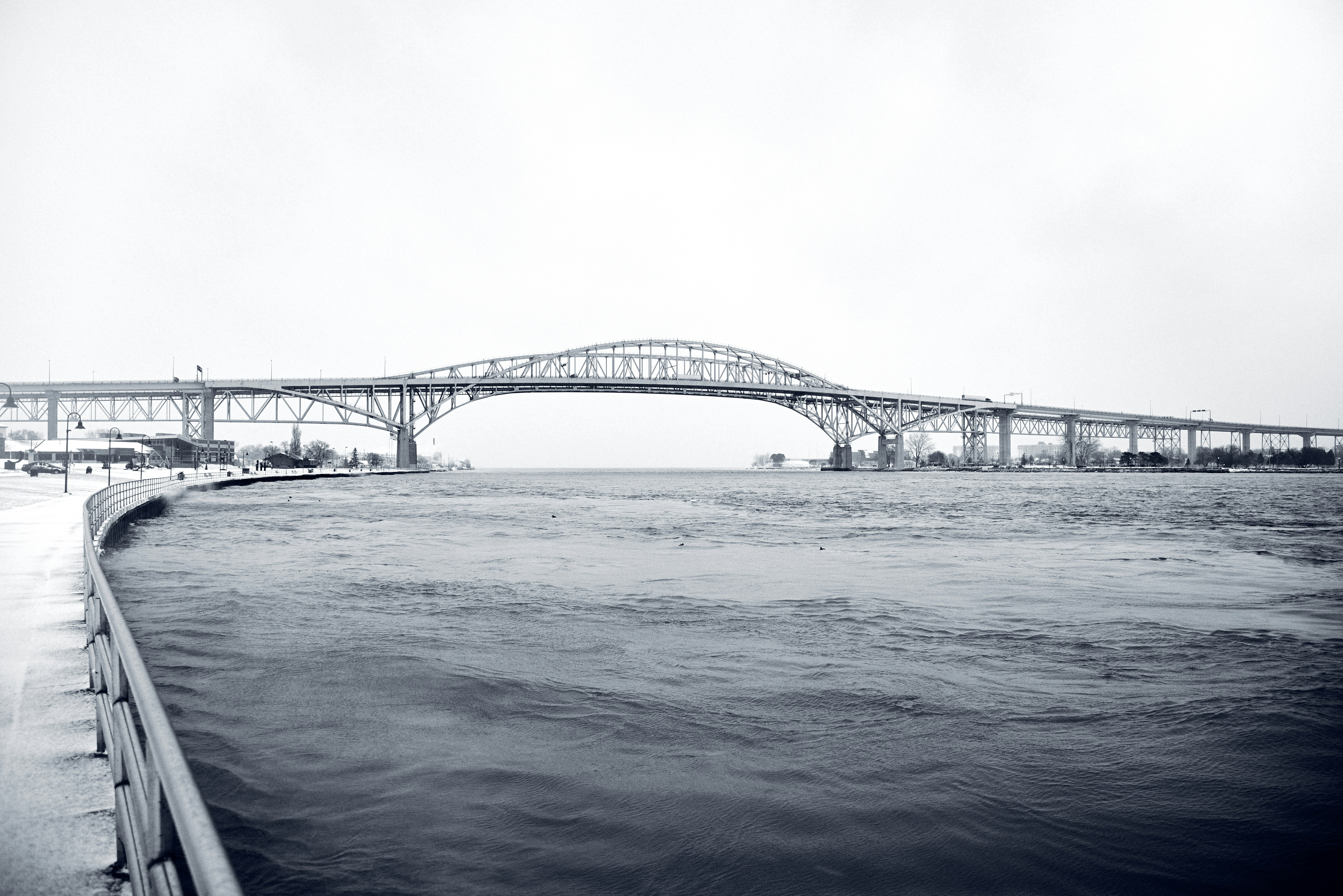 A monochromatic view of a large bridge arching over a calm river, with a winding path along the water's edge.