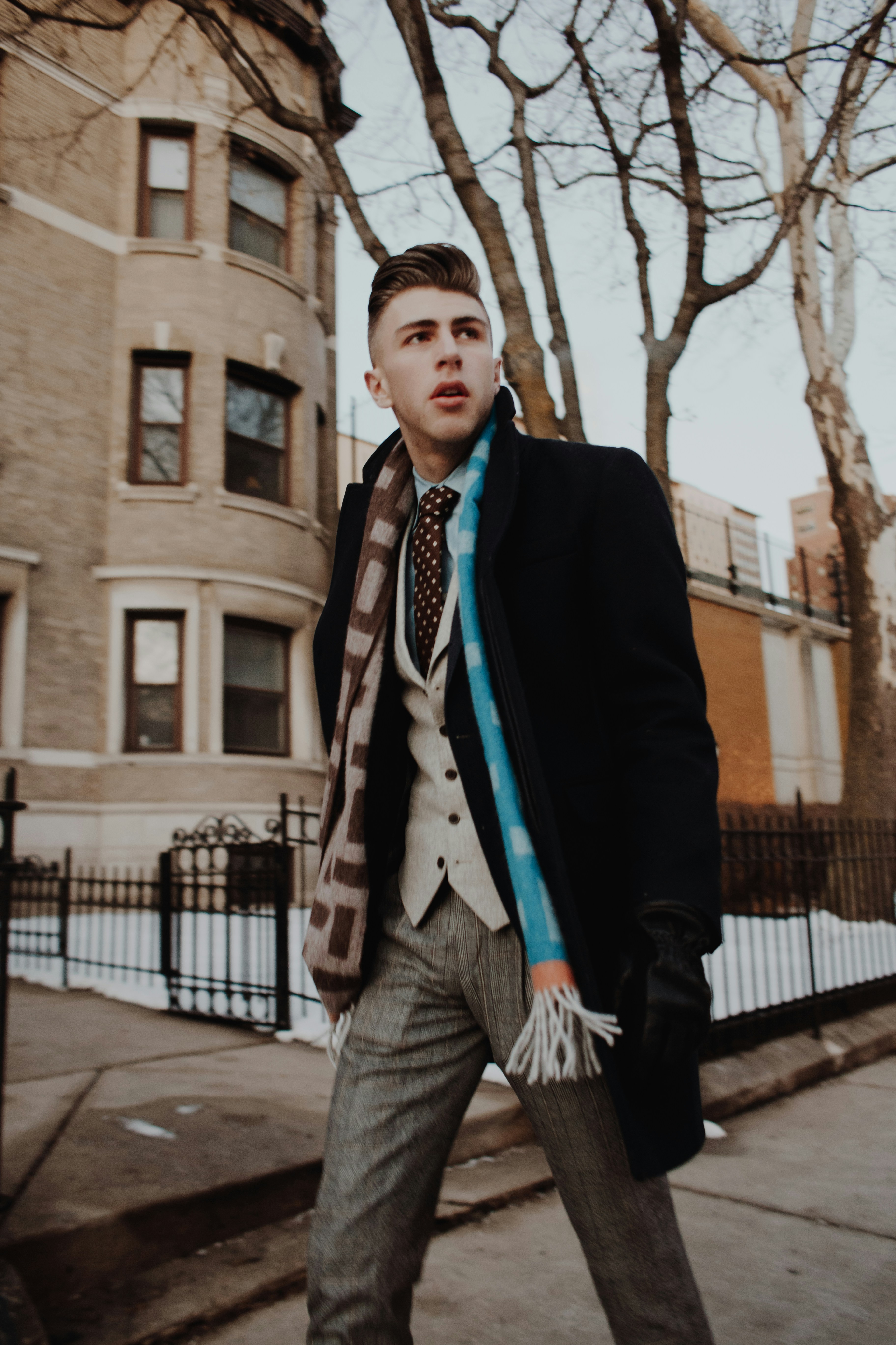 Fashionable young man walking through a city street, showcasing a stylish outfit with a patterned scarf and tailored coat. Urban architecture in the background adds context.