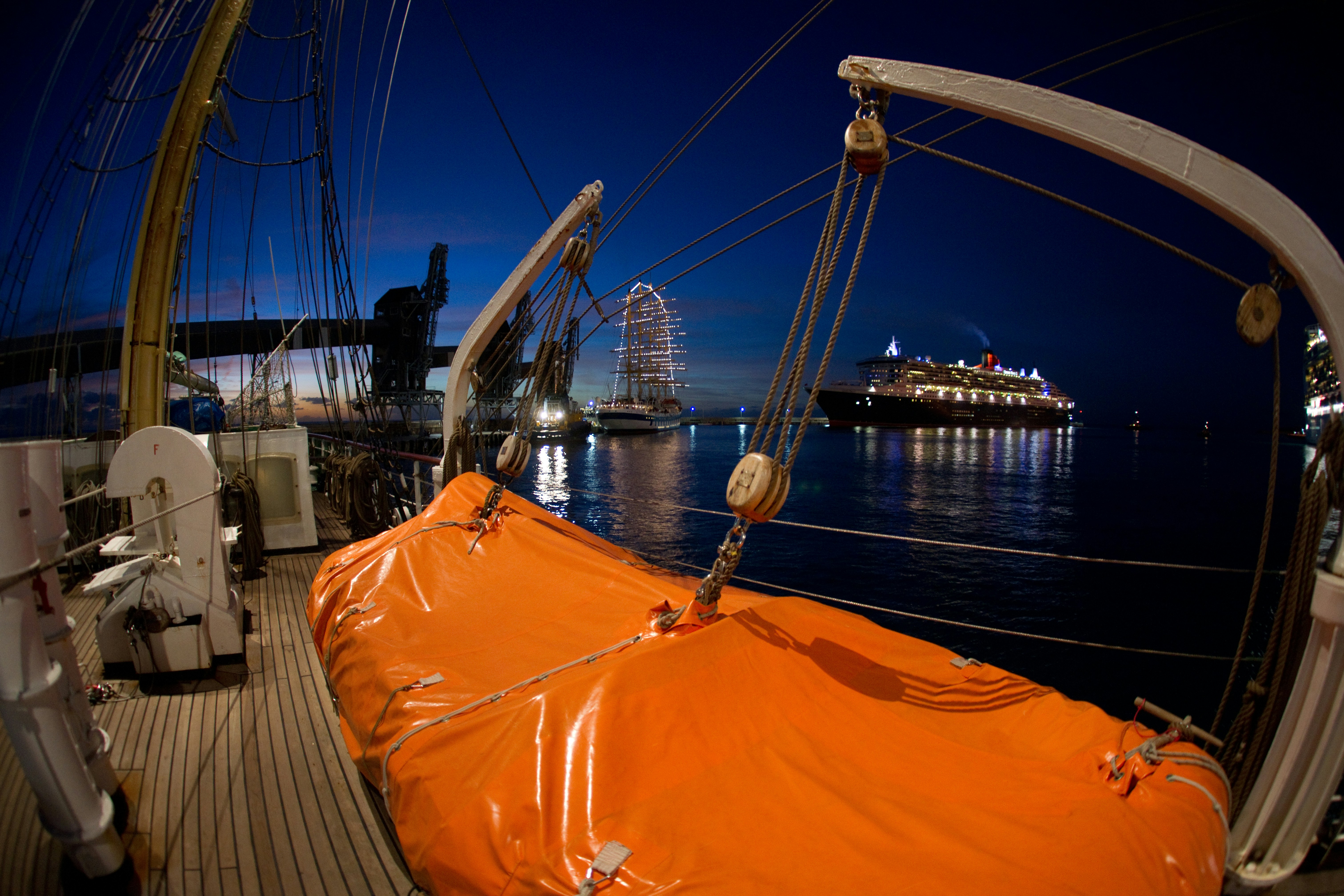 Bright orange lifeboats nestled on a ship's deck with a cruise ship illuminated against the deep blue night sky.