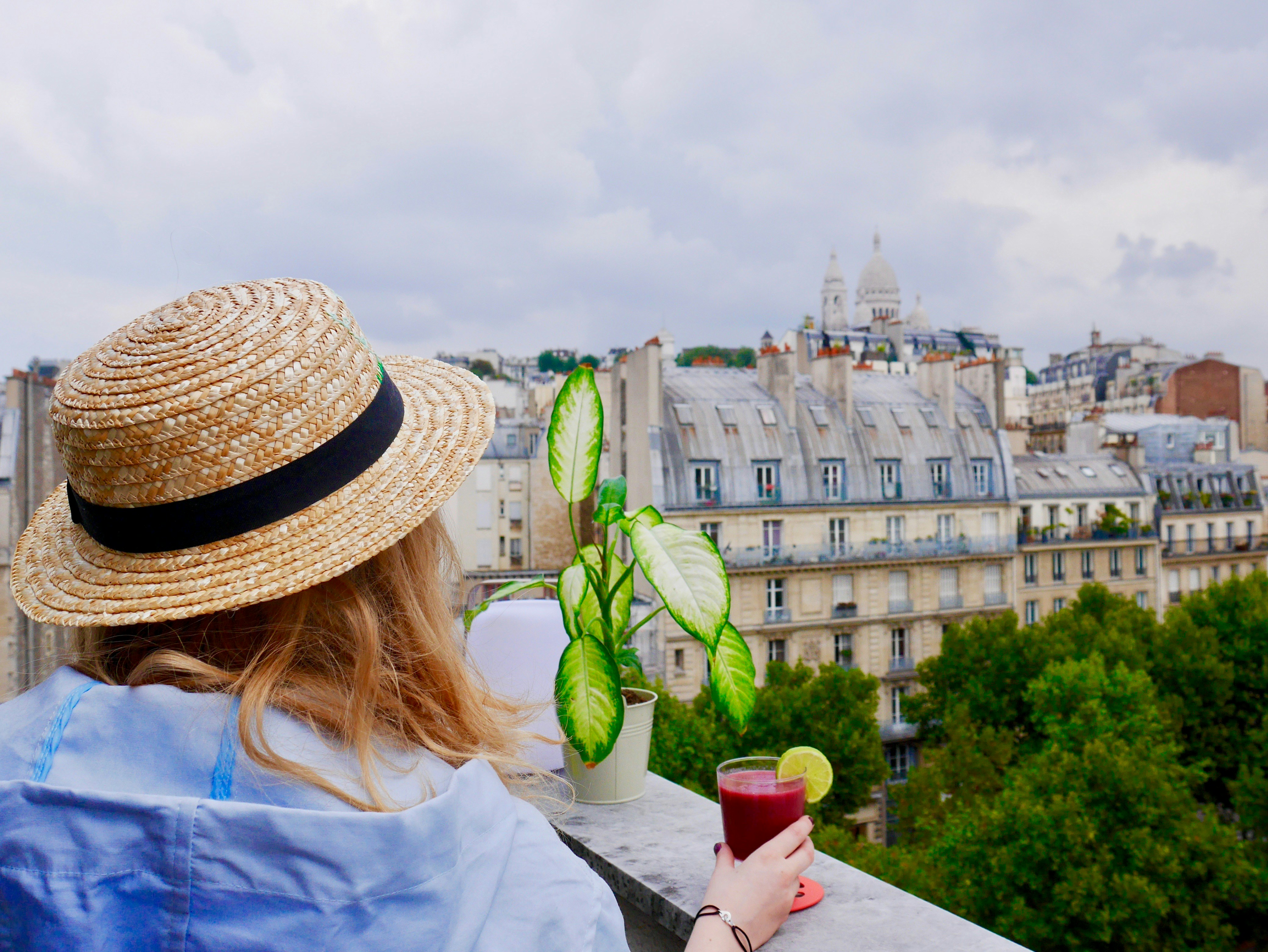 Woman in straw hat enjoys a drink on a rooftop with a view of Sacré-Cœur in the distance.