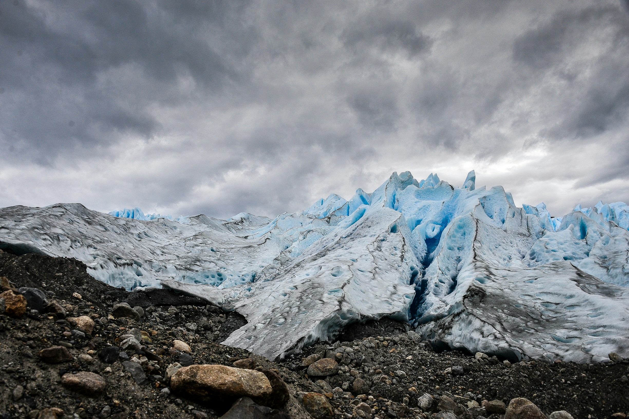 Blue glacier set against a dramatic cloudy sky with rocky foreground.