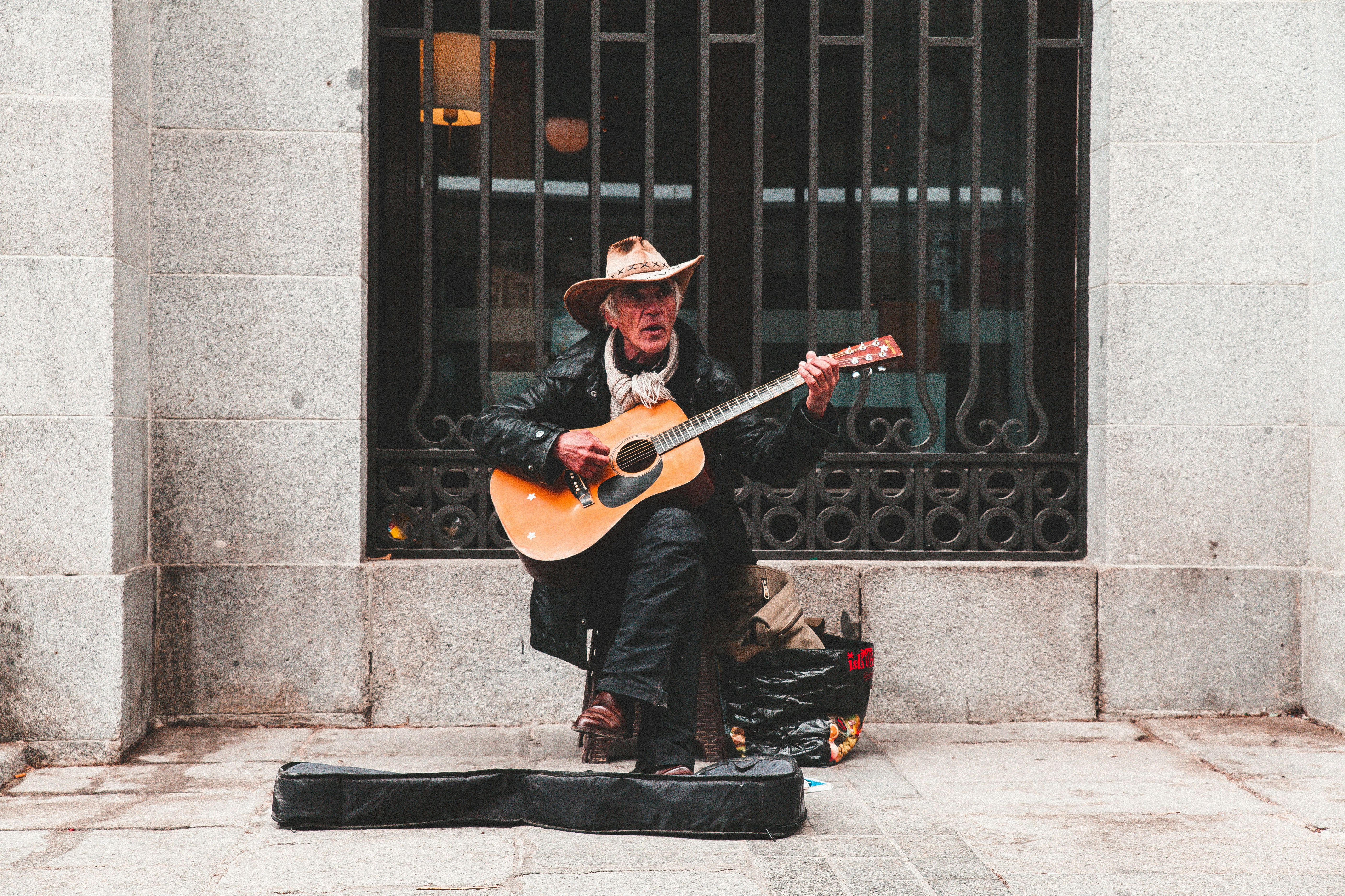 Street musician playing acoustic guitar outside a metal-grated window.