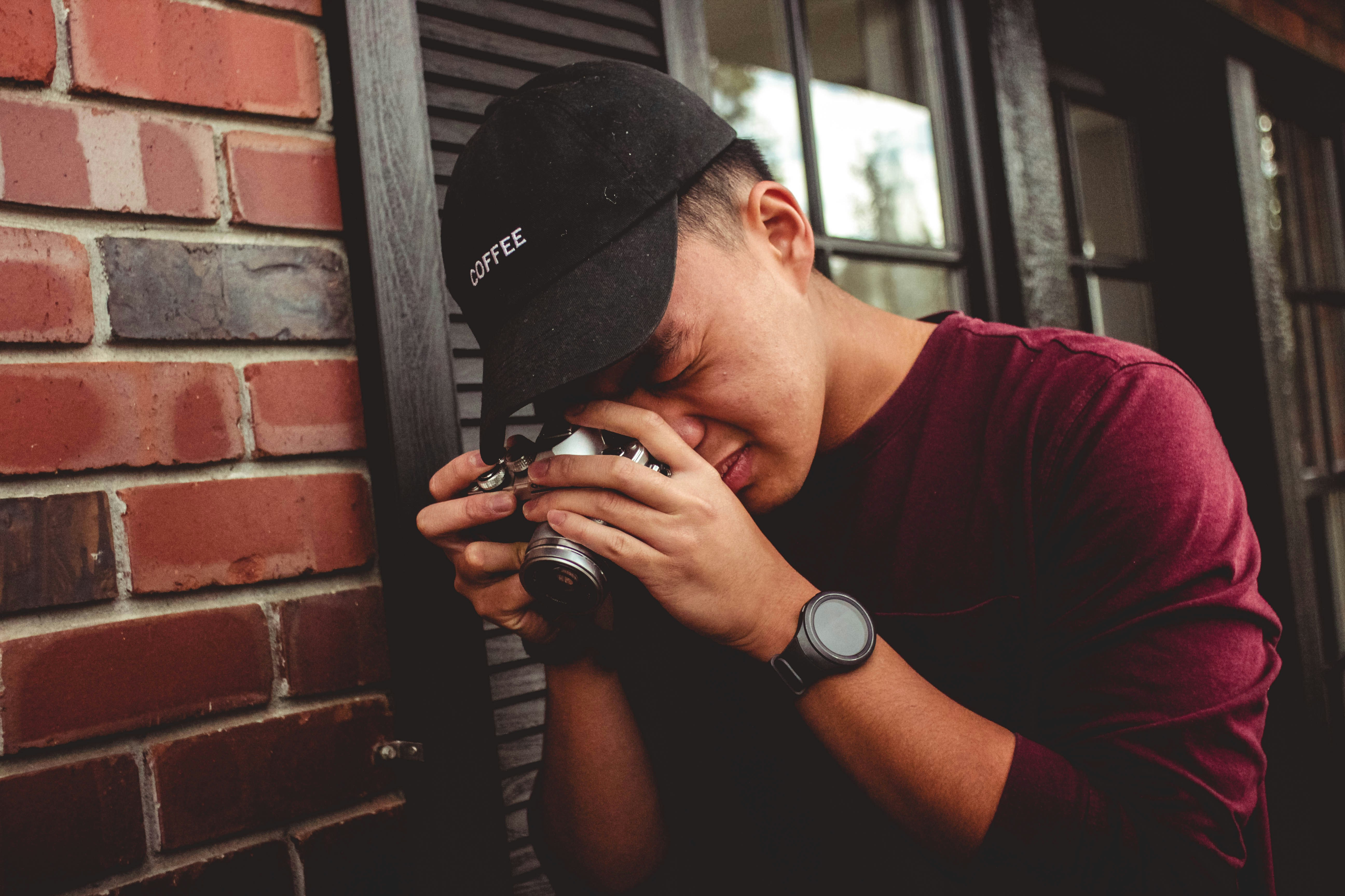 Person in a dark cap using a camera by a brick wall and window.