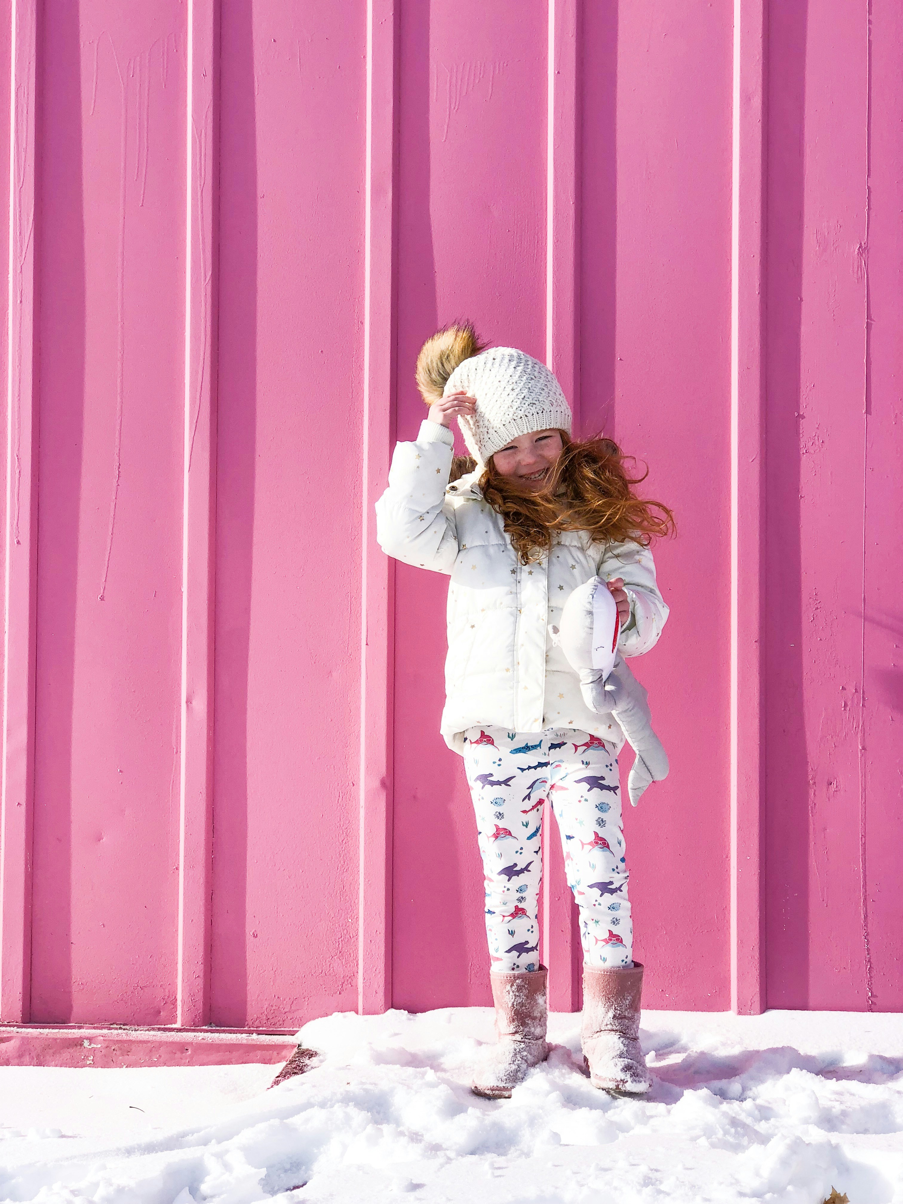 Child in a cozy white jacket and knitted hat poses playfully in front of a vibrant pink wall, surrounded by snow.