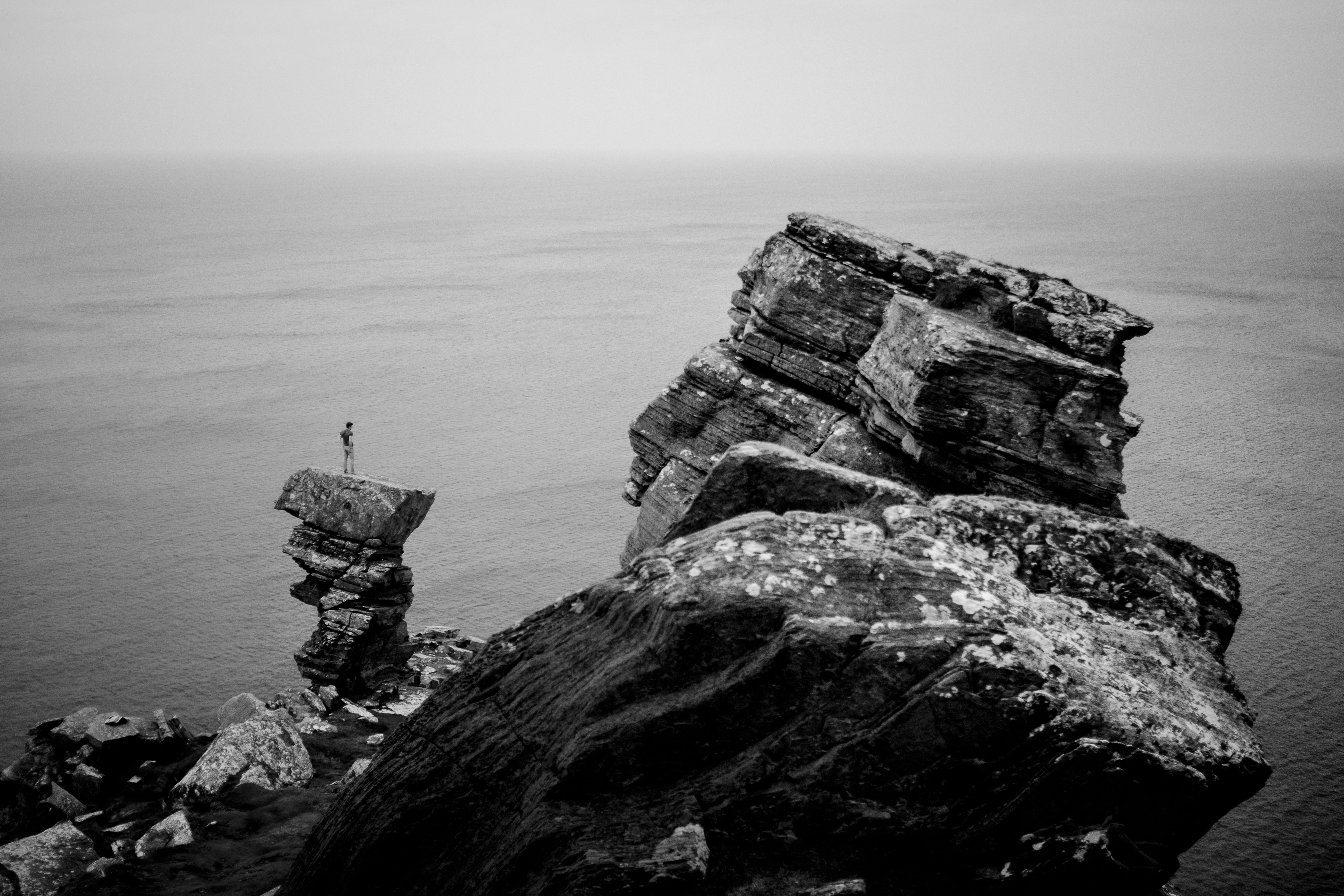 Grayscale photography of a person standing on cliff near the ocean ...