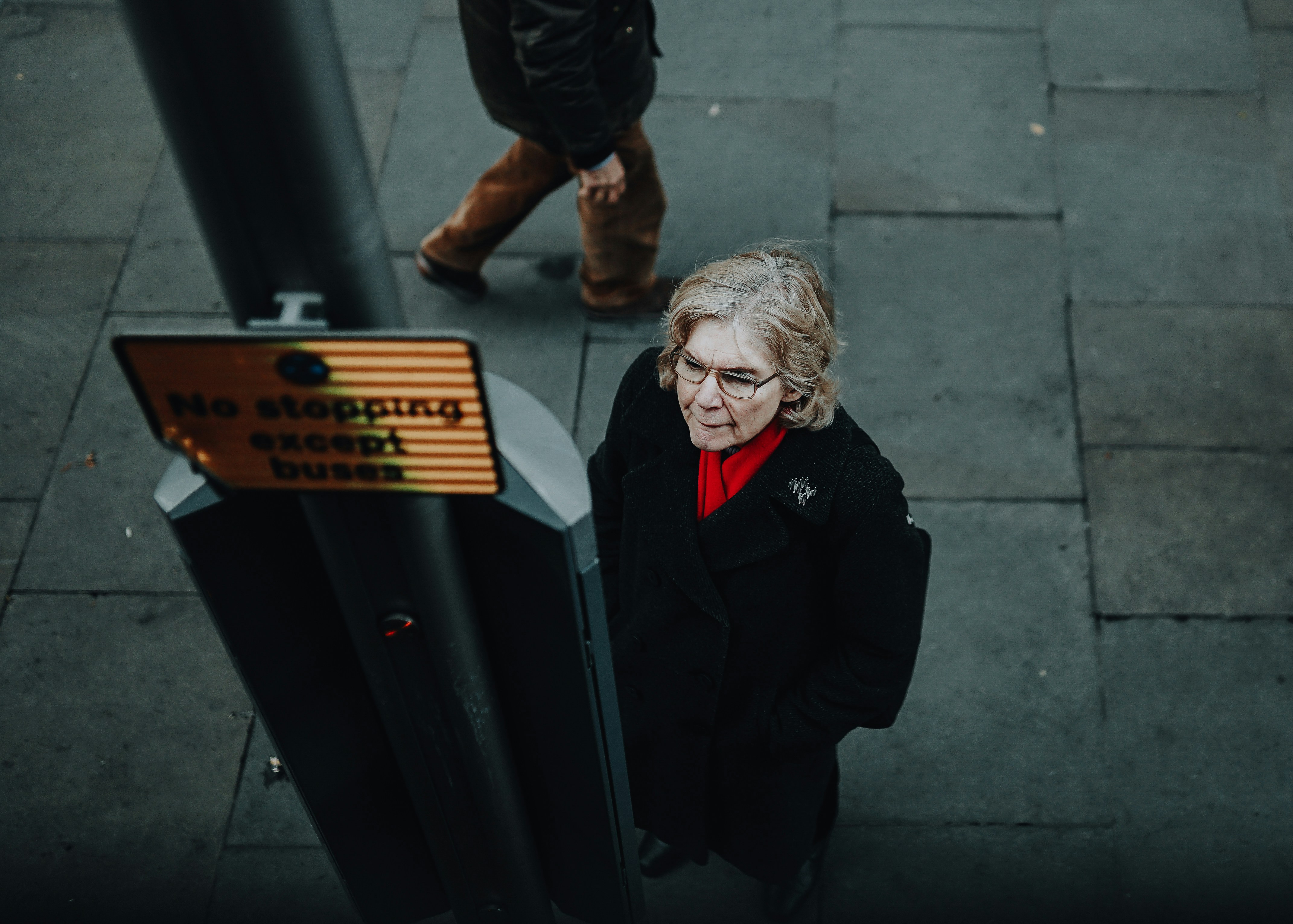 Elderly woman in a black coat with a red scarf stands by a bus stop, gazing thoughtfully. A blurred figure walks past in the background.