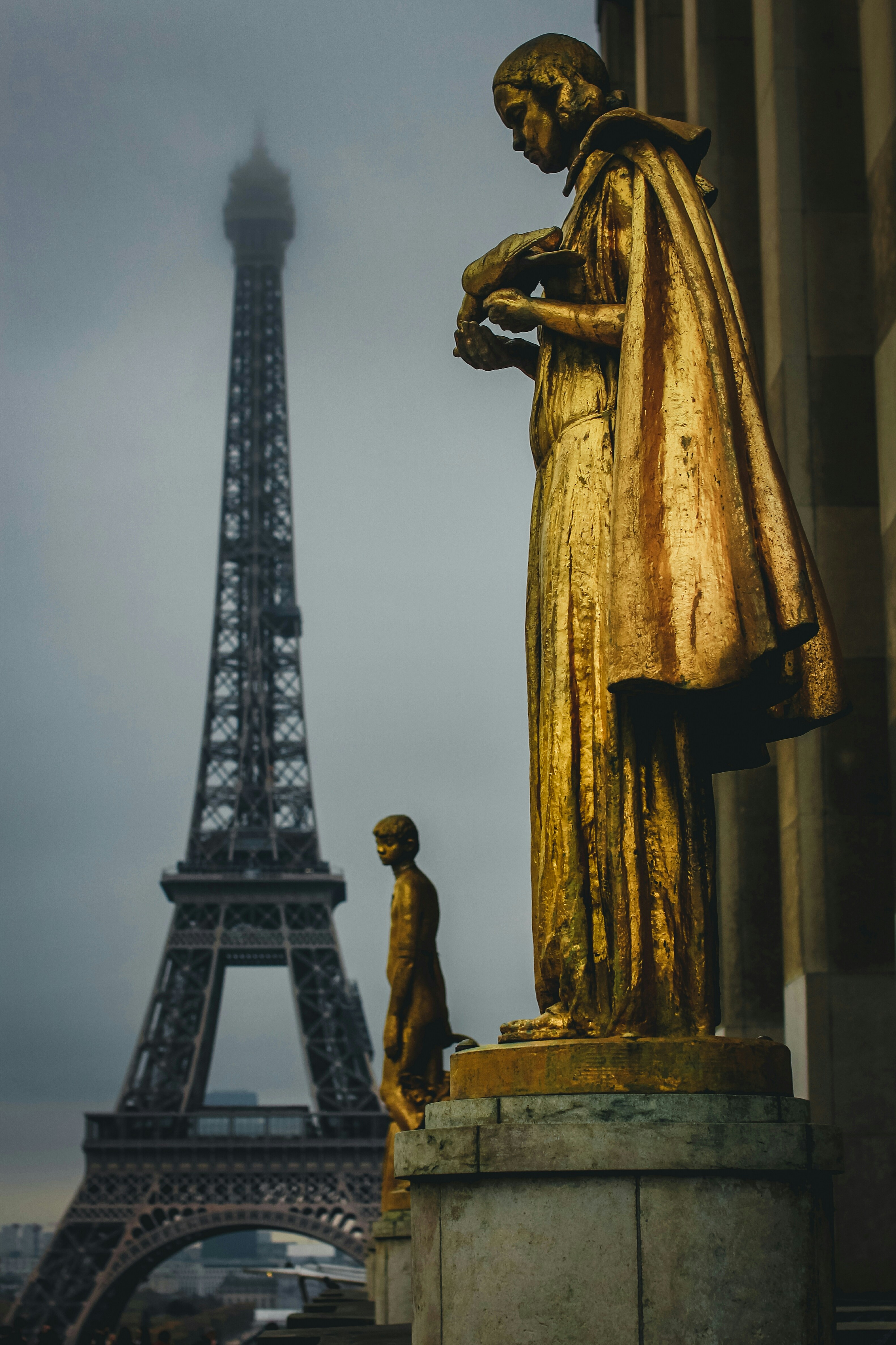 Two golden statues stand watch, framing the Eiffel Tower in the background under a moody sky.