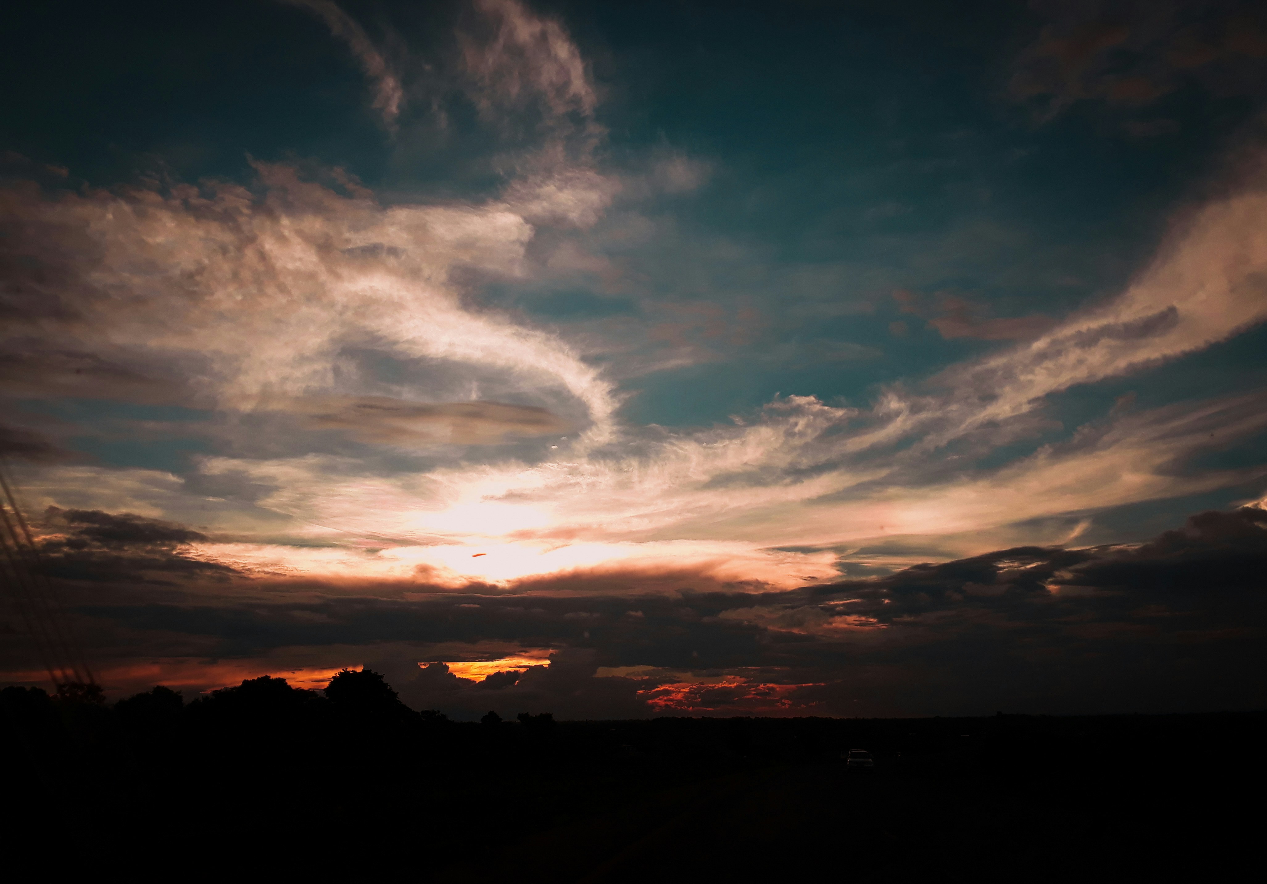 Dramatic clouds swirl in a vibrant sunset sky over a darkened landscape.