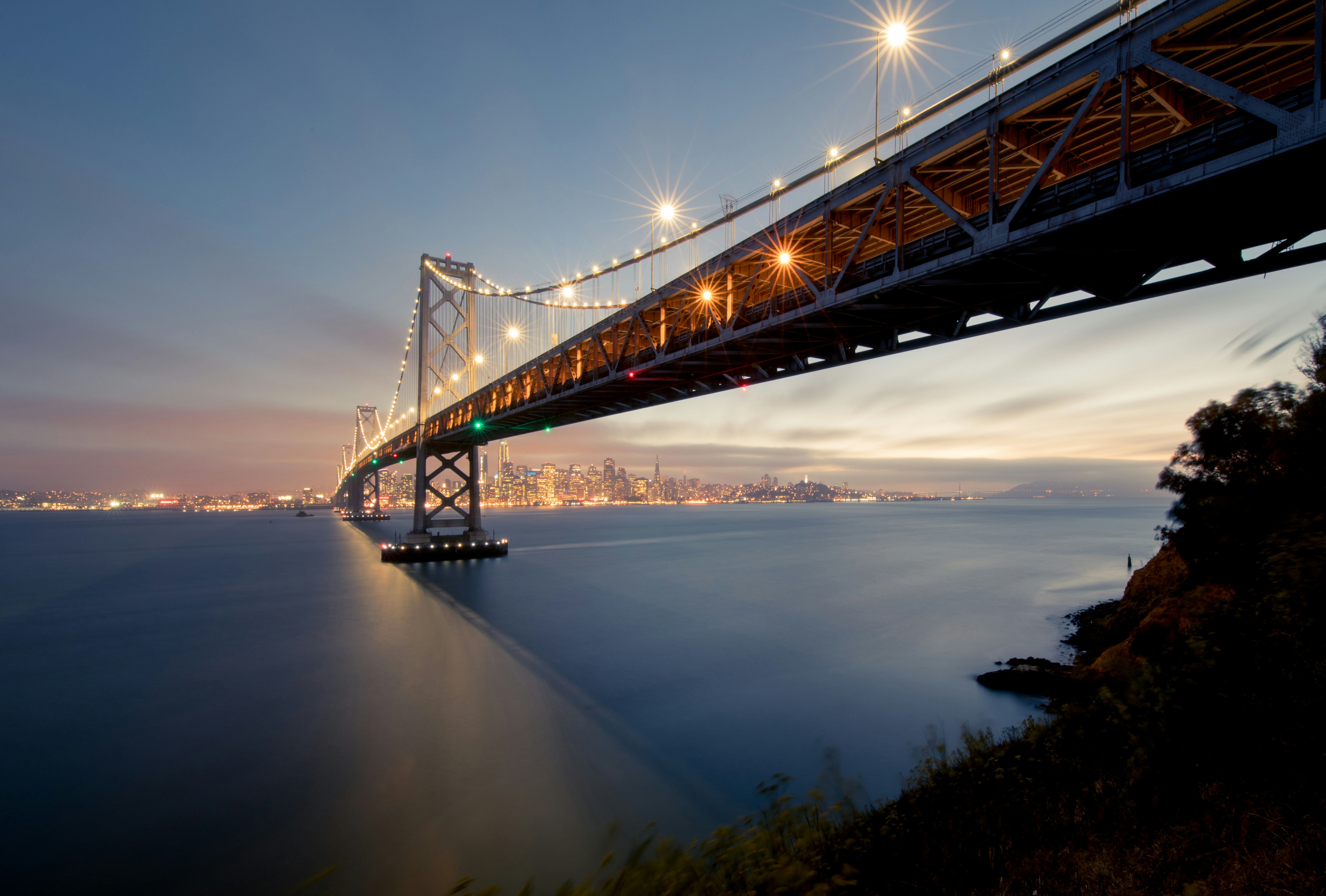 Bay Bridge illuminated against a twilight cityscape with smooth water reflections.