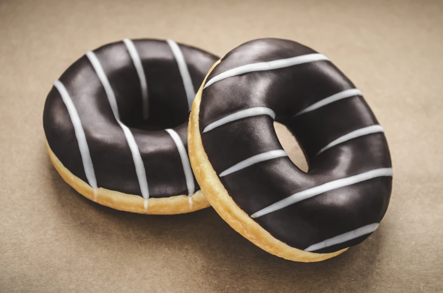 Two chocolate-frosted ring donuts with white drizzle sit on a plain brown paper background. The front donut overlaps the other, creating a simple bakery product composition.