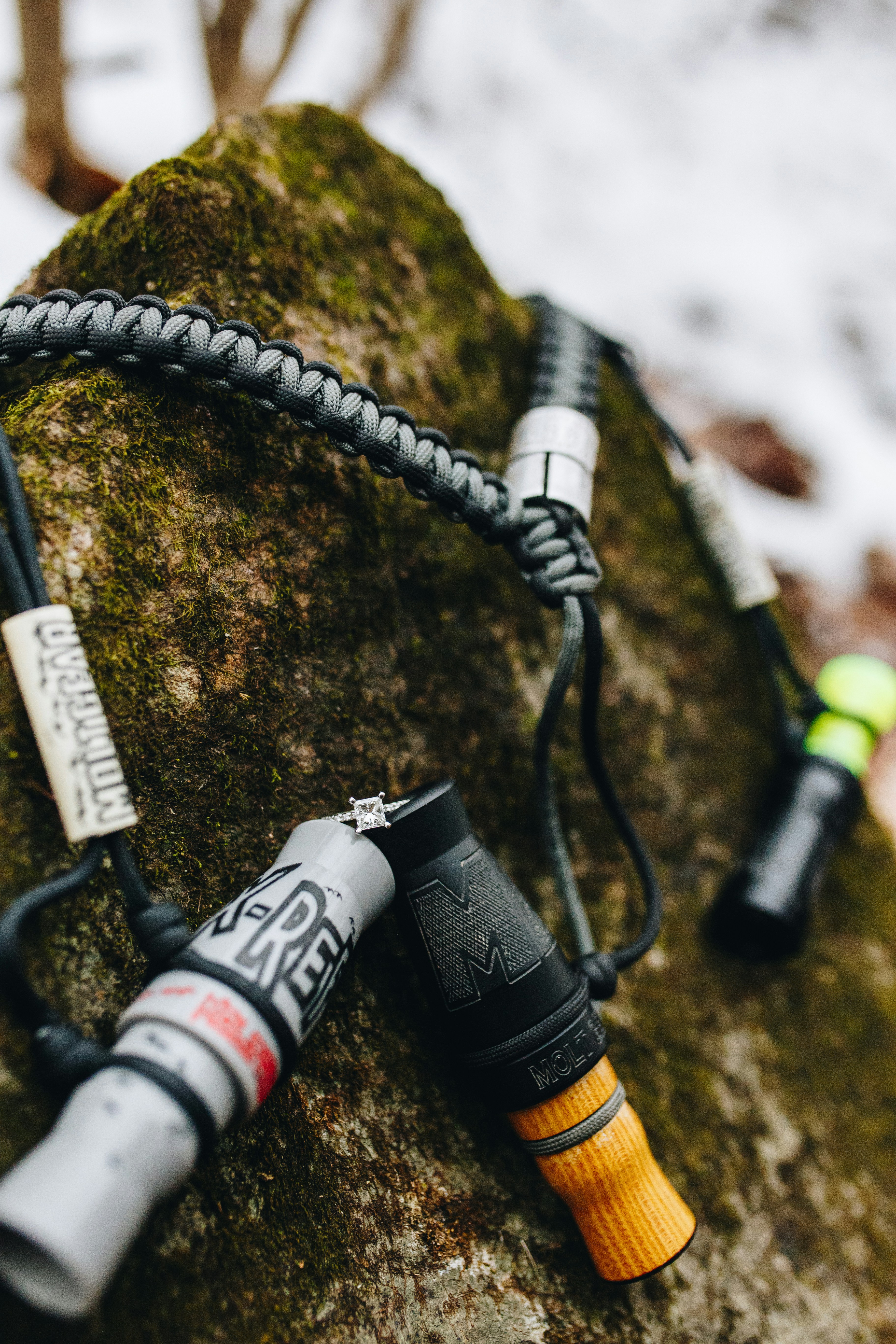 Close-up of braided paracord lanyards and metal hardware resting on a moss-covered rock. Shallow depth of field emphasizes texture and metallic details against a soft, natural background.
