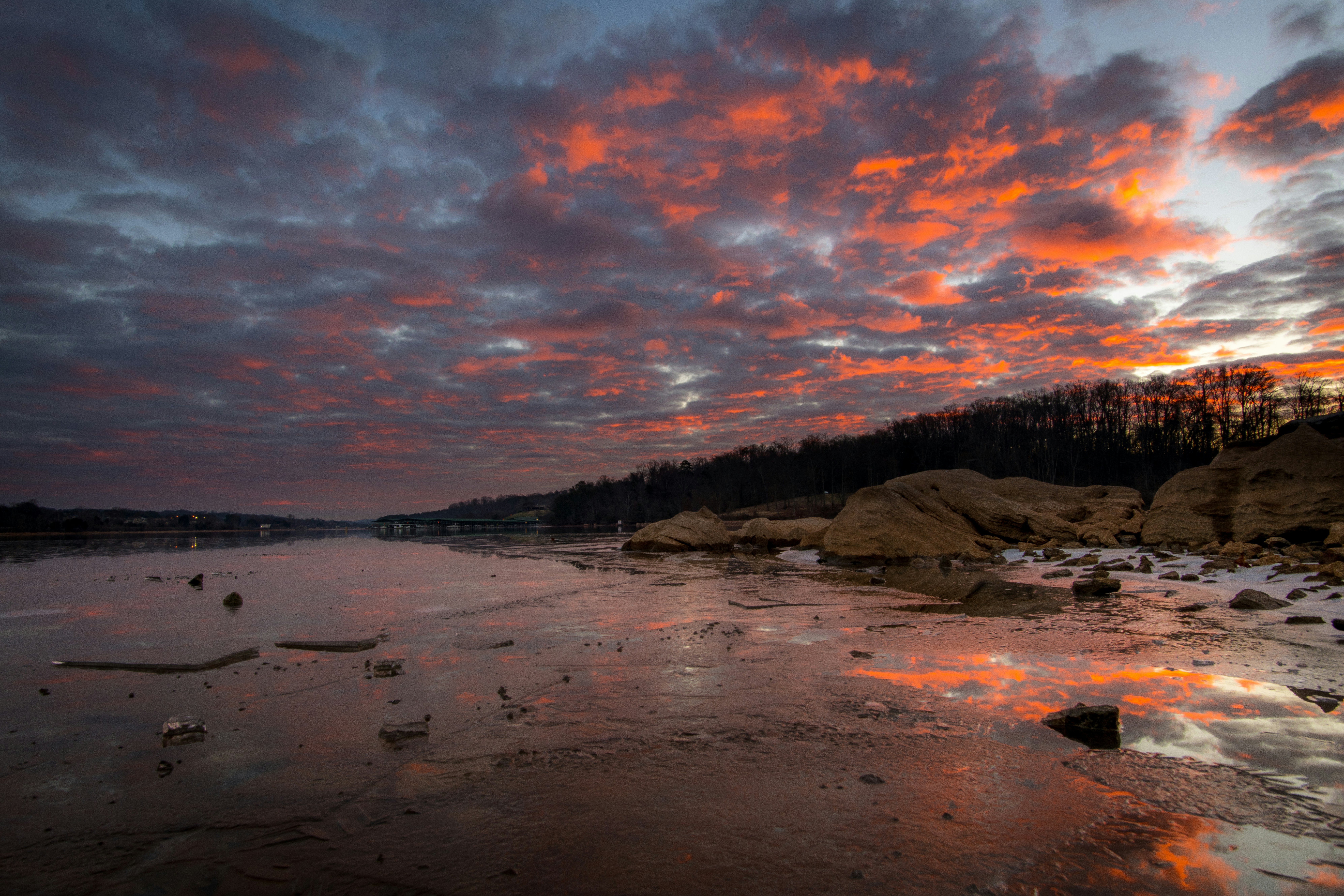 Vibrant sunset reflecting on a tranquil shoreline with scattered rocks and dramatic clouds.