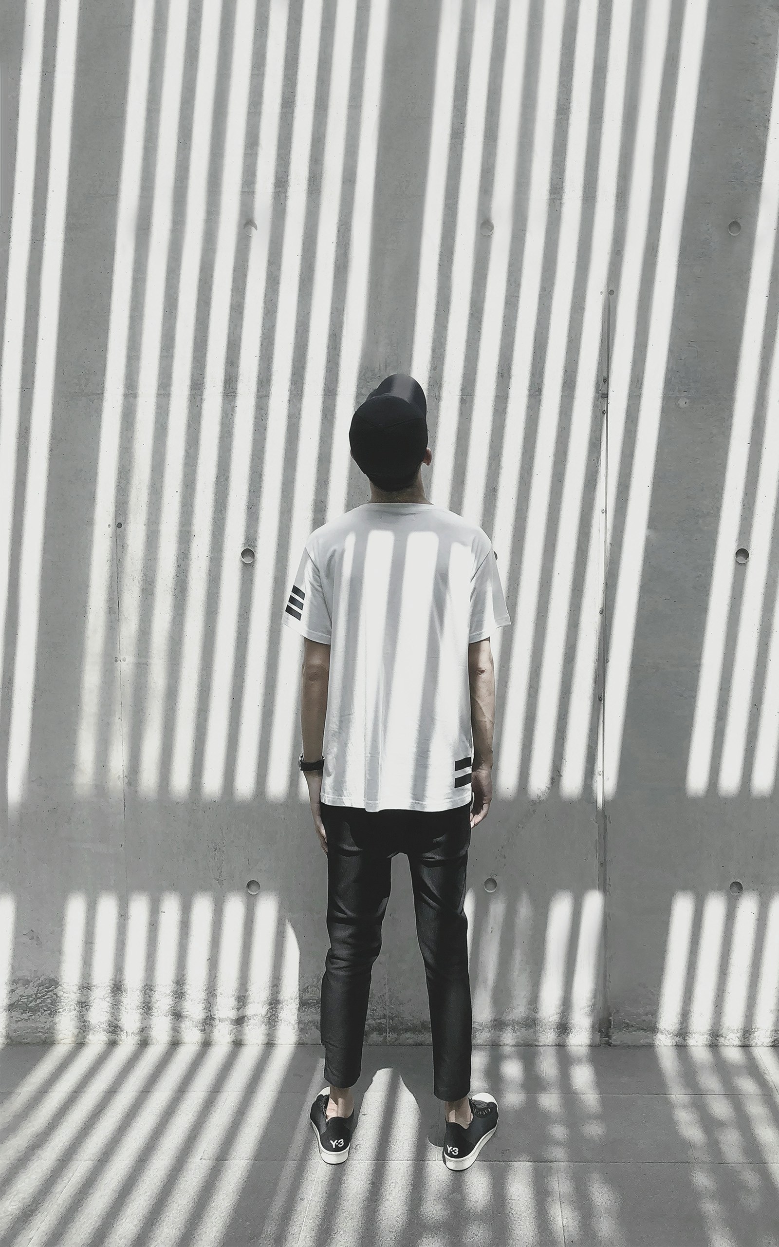 A person stands in a minimalist space, illuminated by stark shadows created by vertical slats, enhancing the contrast between light and dark.