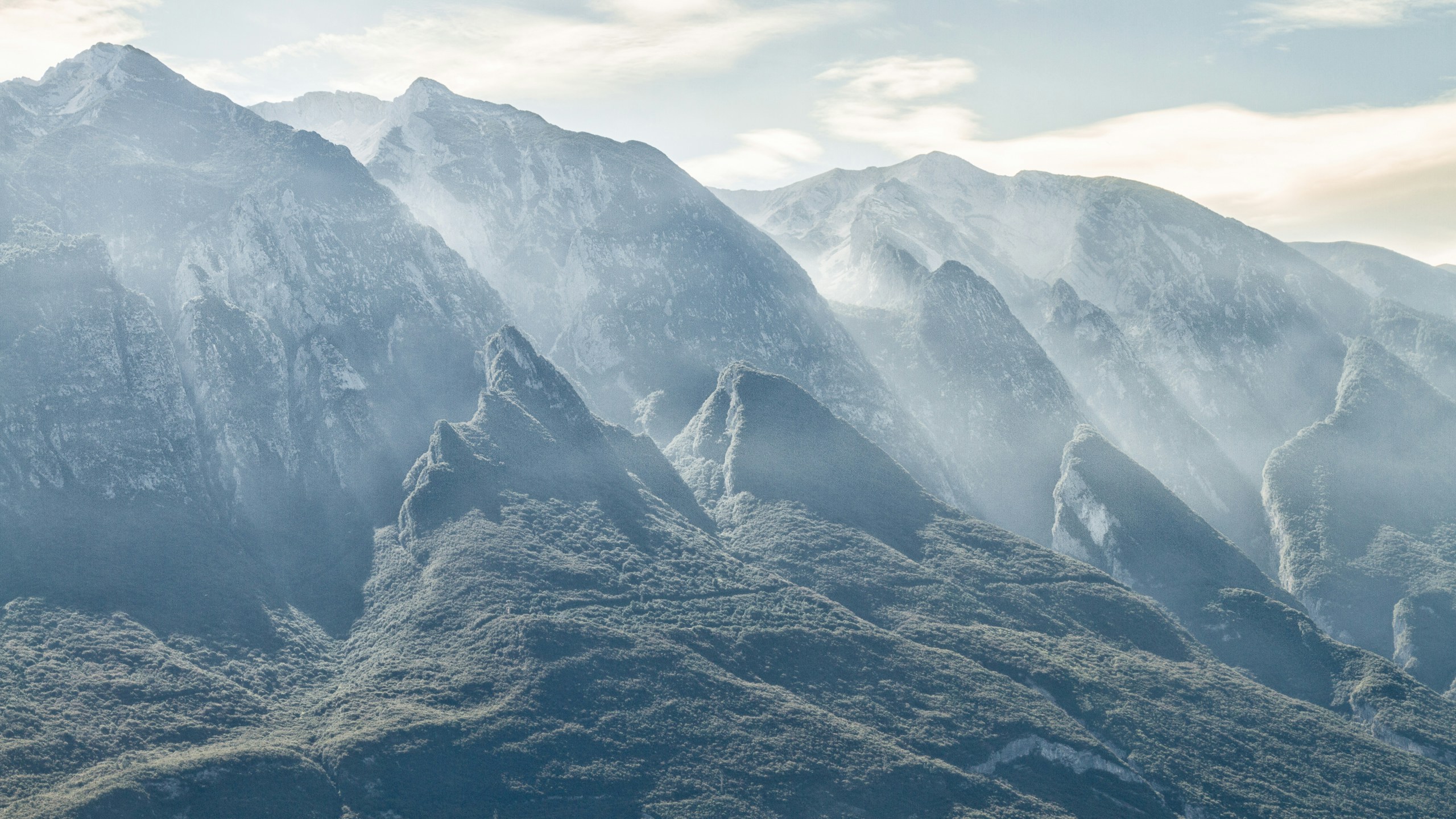 Majestic mountain range shrouded in mist, showcasing jagged peaks and lush greenery beneath a soft sky.