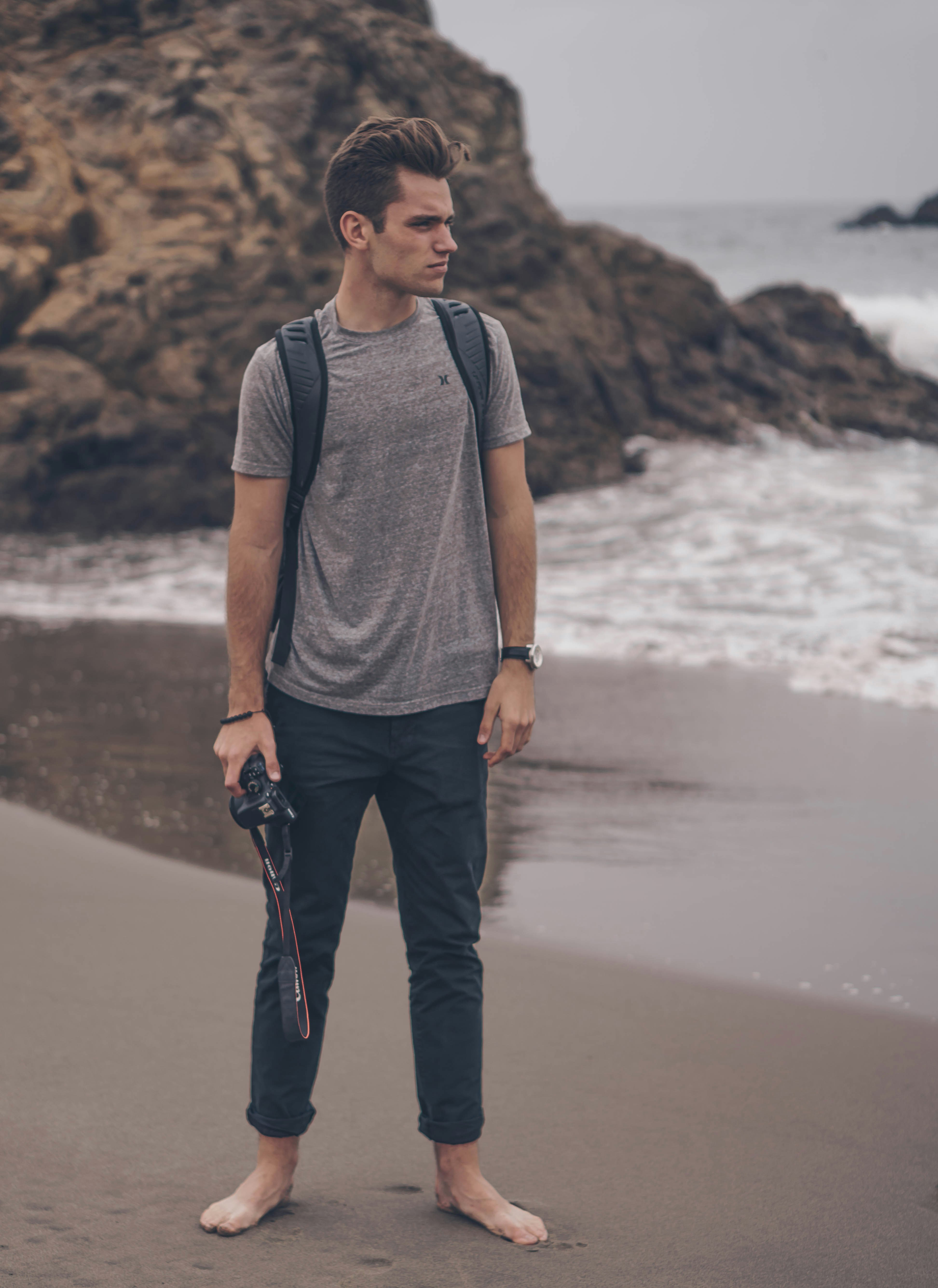 Young man standing barefoot on sandy beach, holding a camera, with rocky coastline and ocean waves in the background.