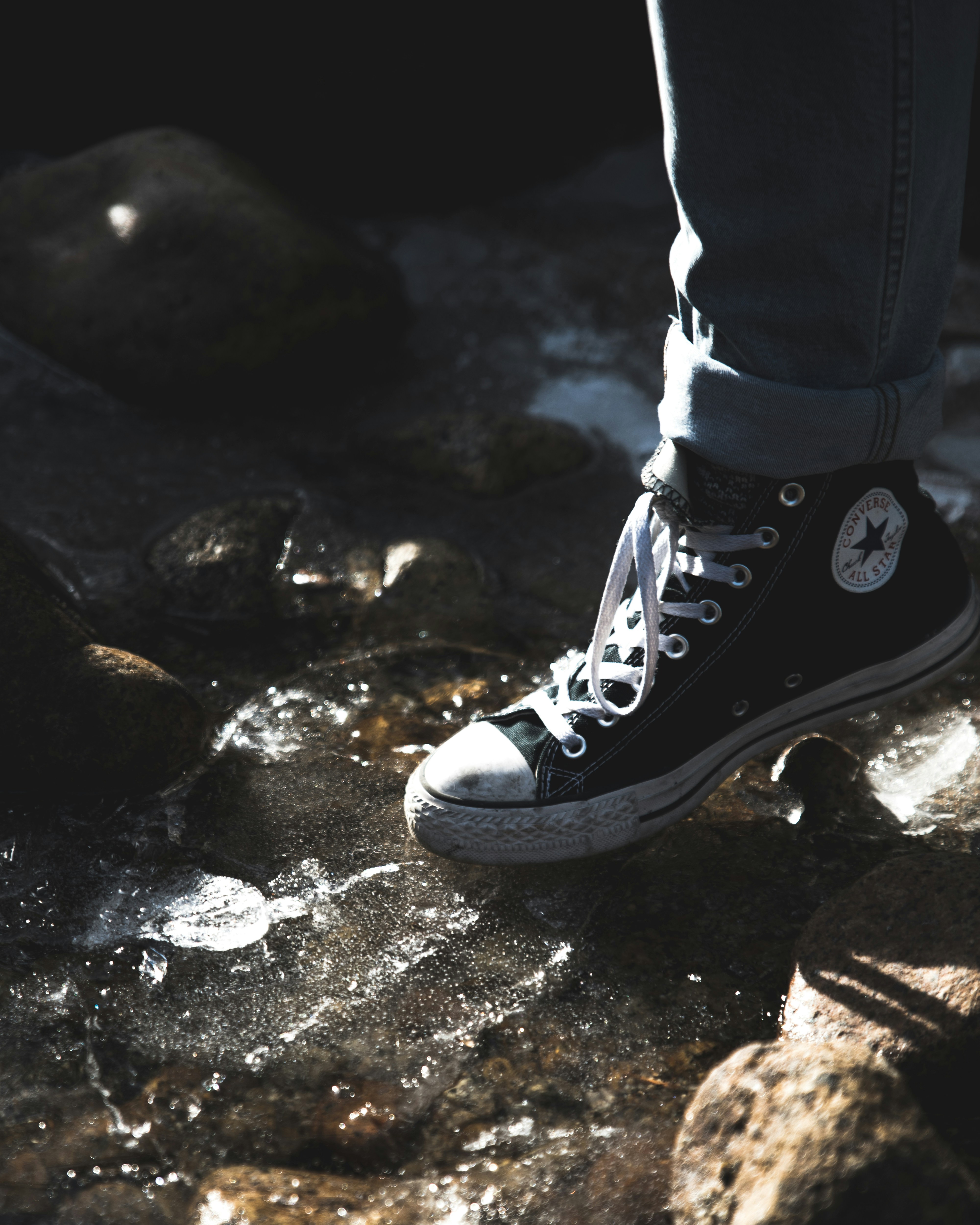 Black Converse sneaker standing on a rocky surface with shimmering water reflections. The scene captures a moment of exploration in a natural setting.