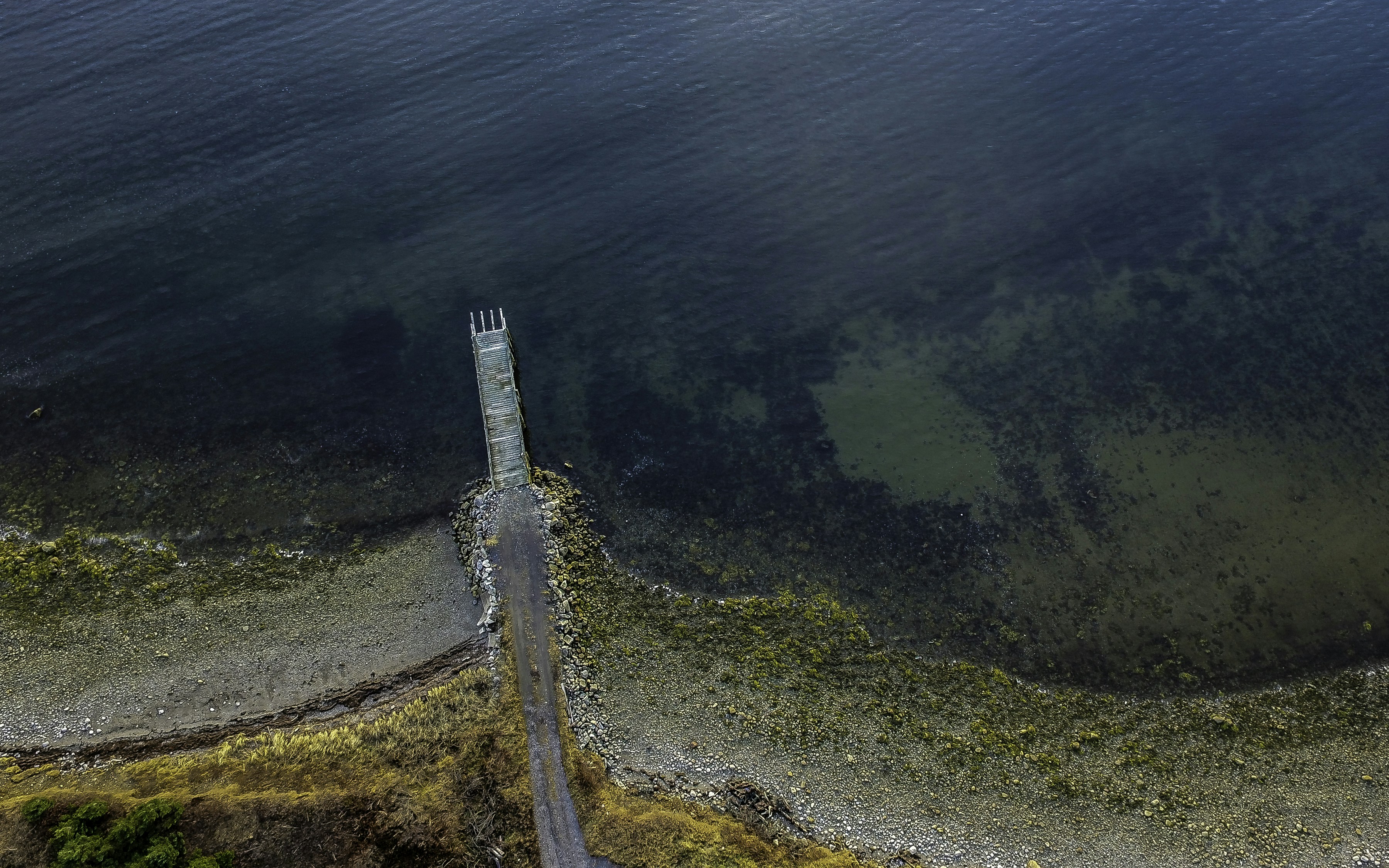 Aerial view of a wooden pier extending into calm waters, surrounded by a rocky shoreline and patches of greenery.
