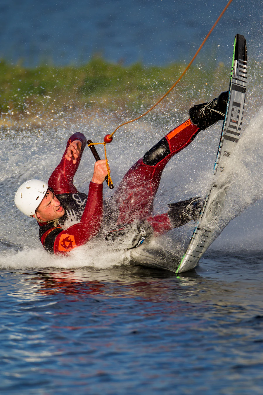 Wakeboarder in red wetsuit and white helmet falling backward while gripping a tow handle as spray explodes around them.