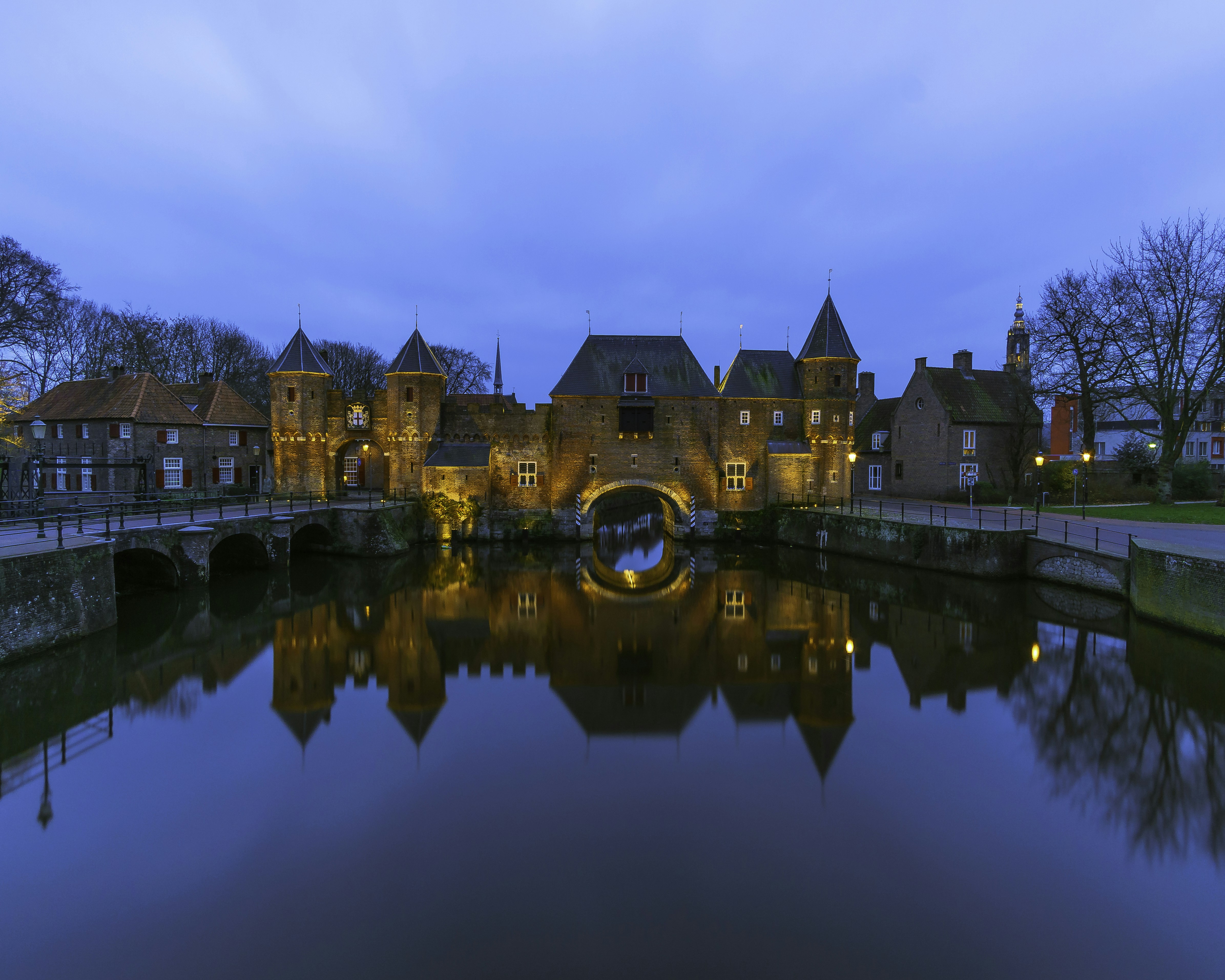 Illuminated medieval fortress and bridge reflecting in calm waters under a dusky blue sky.