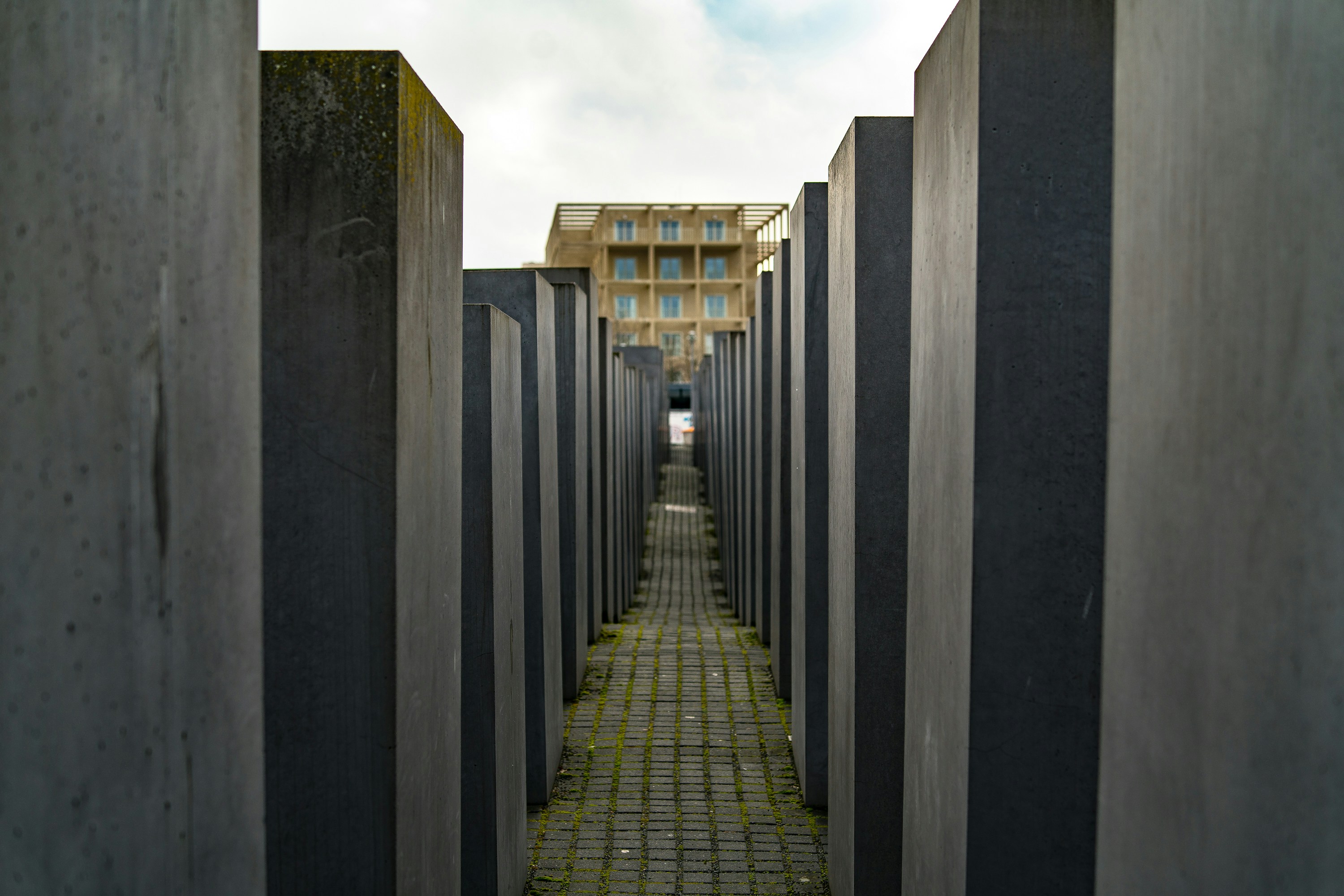 A narrow pathway lined with towering dark pillars, leading towards a distant building, creating a somber yet reflective atmosphere.