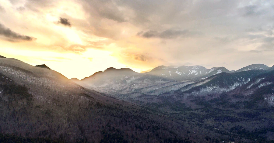 Aerial landscape photograph of snow-dusted mountains at sunset, with warm light brushing the left ridge and a distant forested valley under a moody sky.