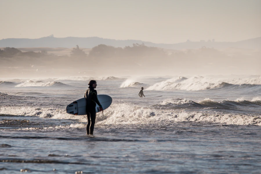 Surfer standing in shallow water with a board, another surfer riding a wave in the distance on a misty morning by the sea.