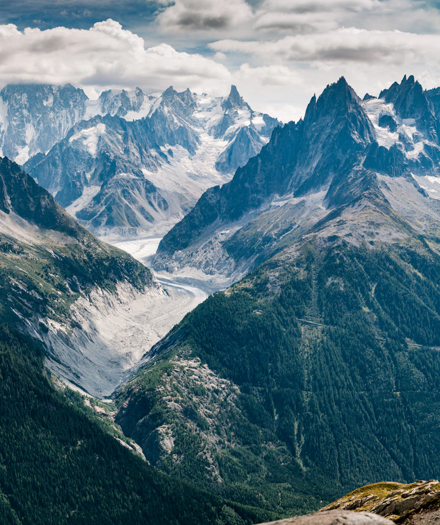 A sweeping mountain range with sharp granite peaks rising above a glacier-carved valley and dark forested slopes. Thick clouds hang over the rugged alpine scene.
