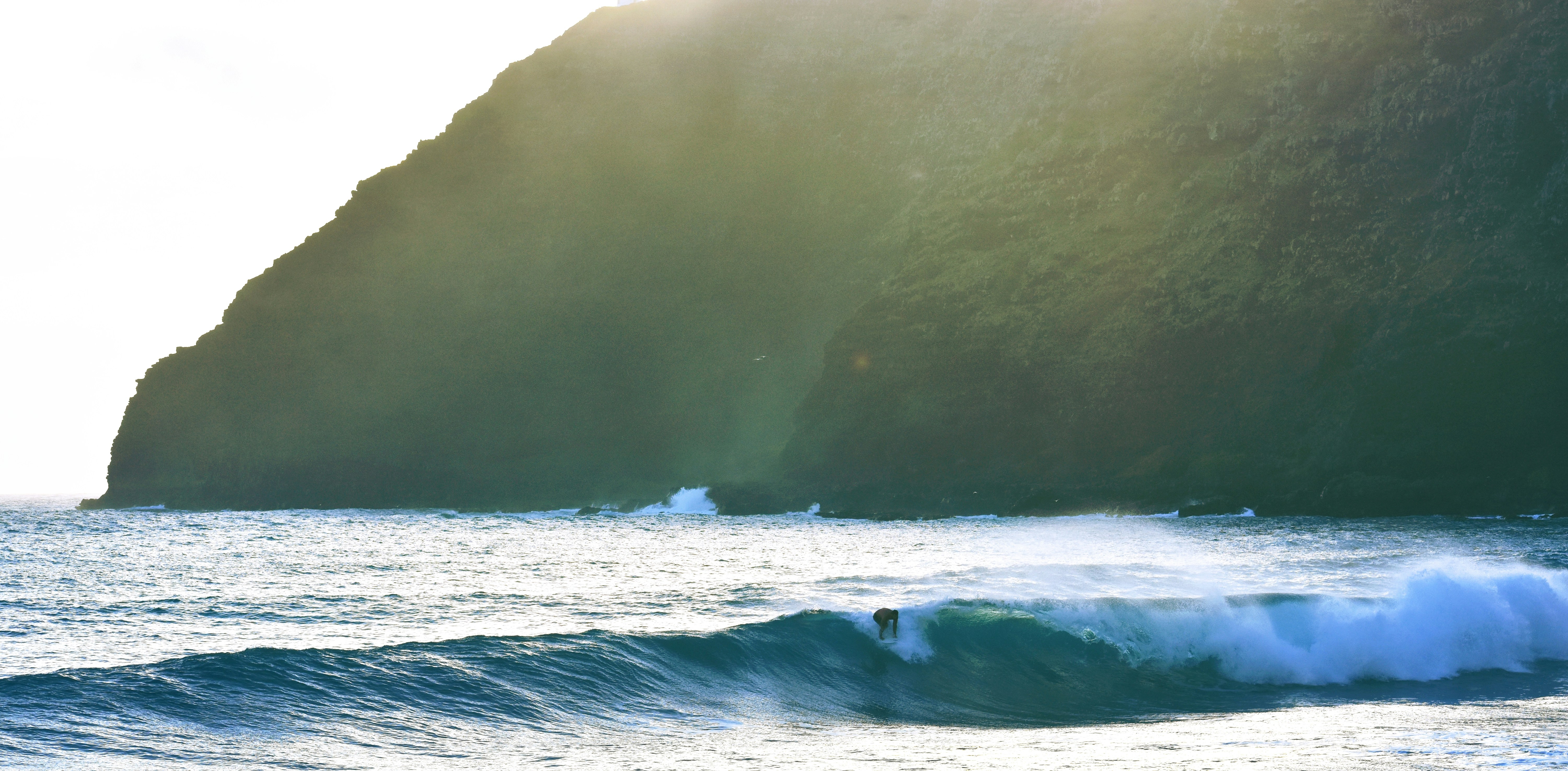 Surfer navigating a wave with a dramatic cliff backdrop under soft, diffused light.