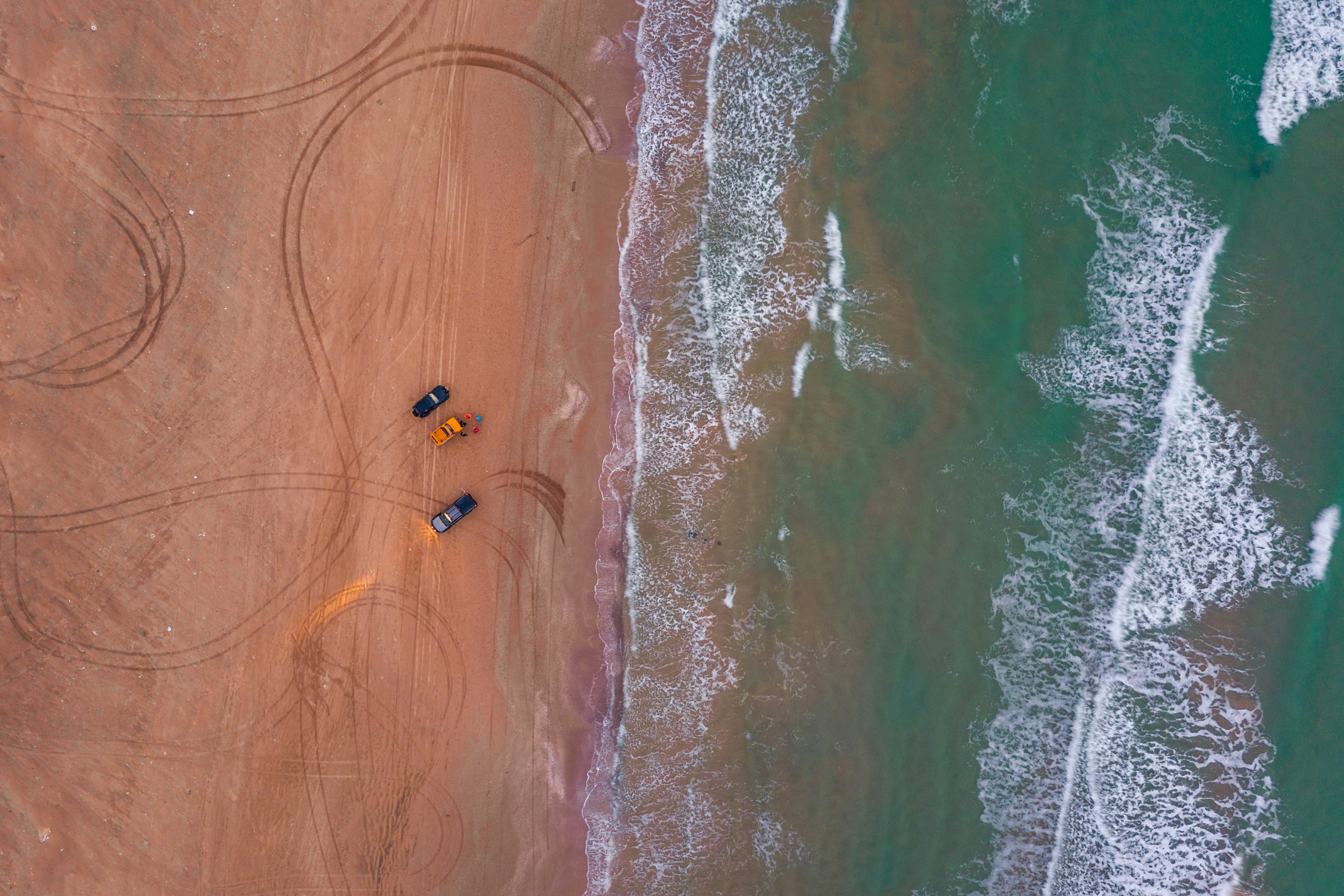 Three vehicles parked on a sandy beach with tire tracks curving towards the turquoise ocean waves.