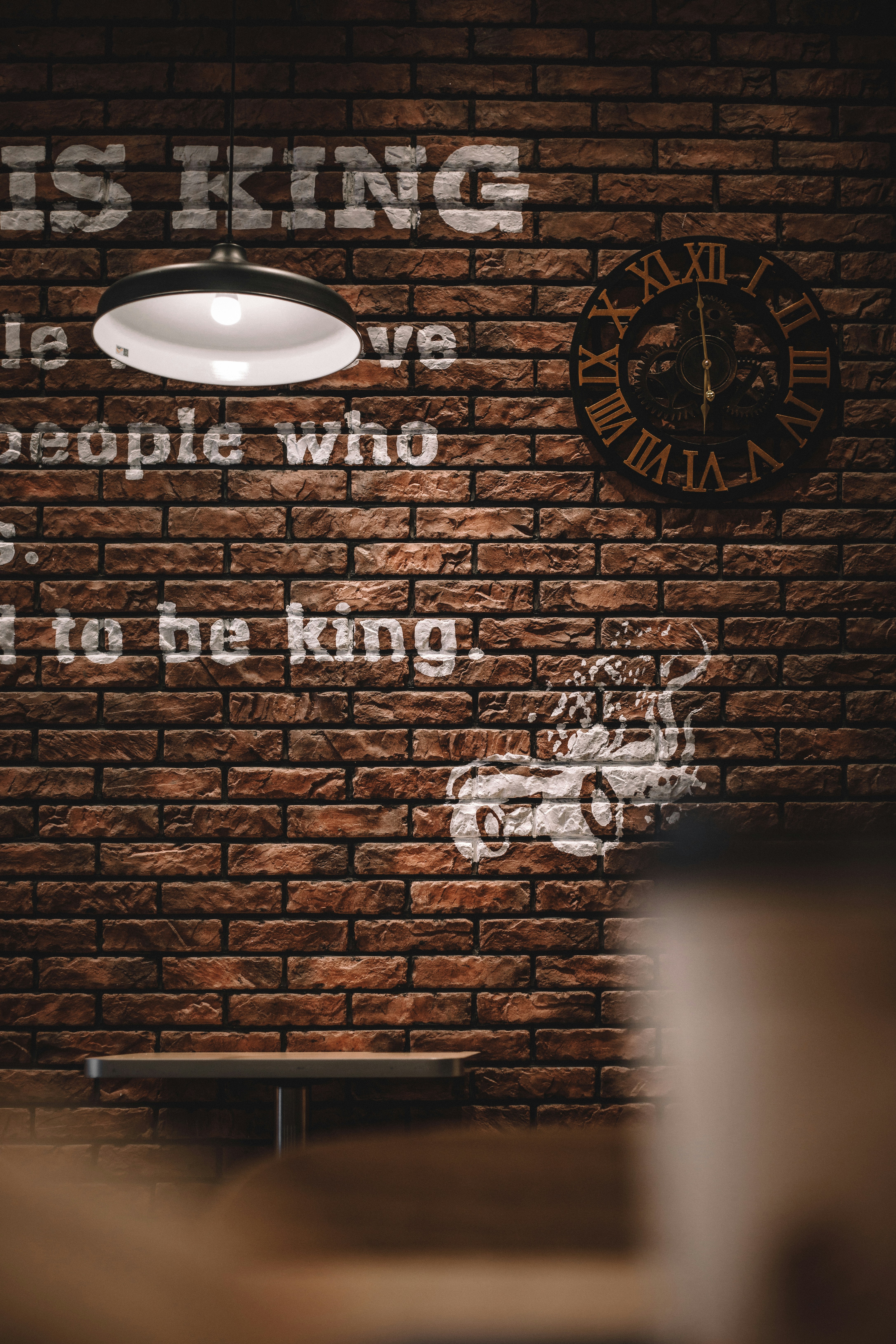 Vintage clock and motivational text on a brick wall in a café, with a blurred foreground hinting at a warm atmosphere.
