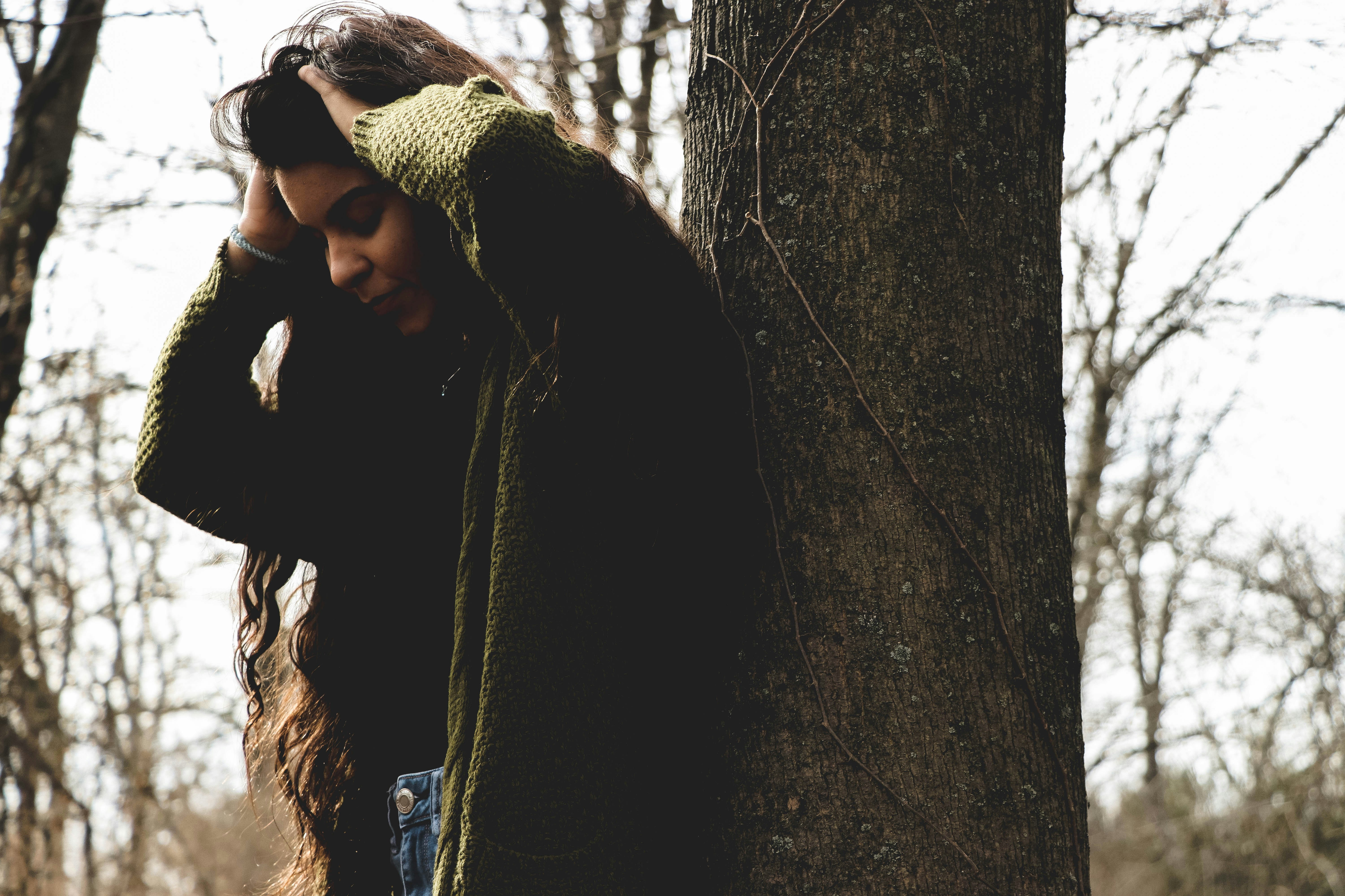 Person leaning against a tree in a forest, hands in hair, surrounded by bare branches.
