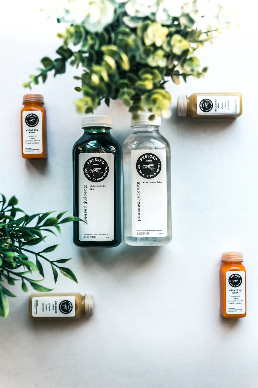 Assorted pressed juice bottles arranged on a light background with leafy greenery around them. Two large bottles sit centered among smaller orange and pale juice shots.