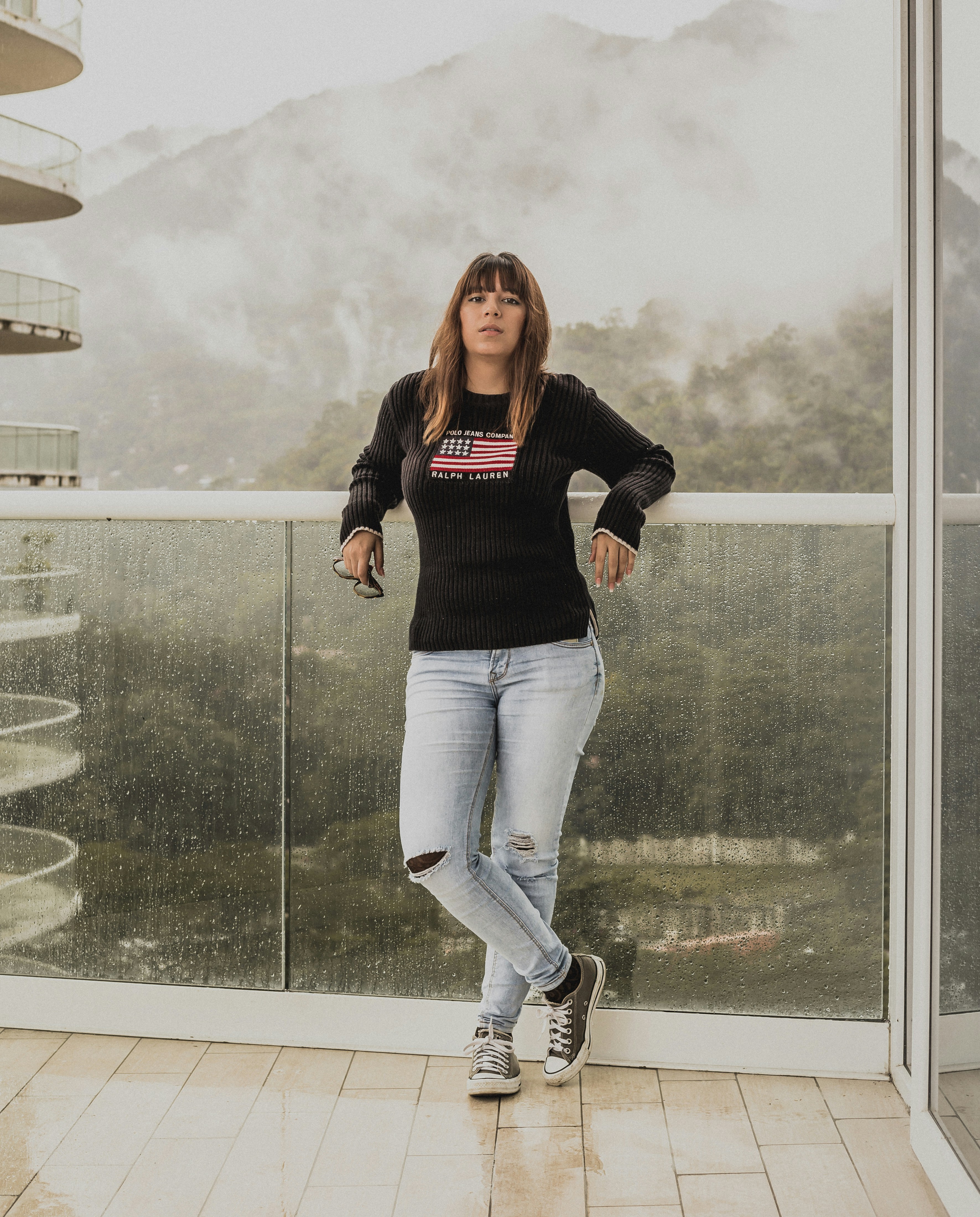Woman in a black sweater with an American flag design leans against a glass railing, overlooking a misty mountain backdrop.
