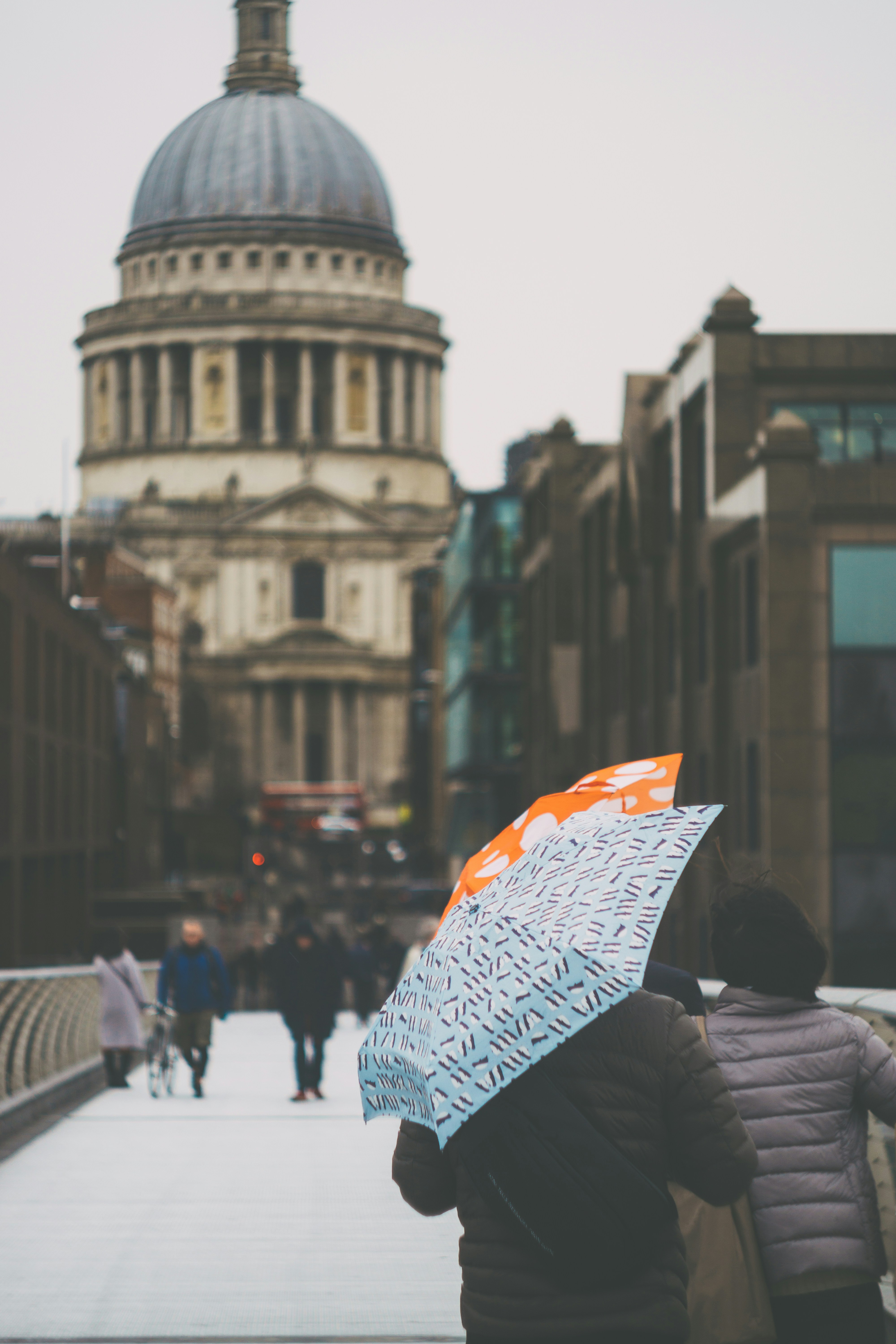 Couple walking with colorful umbrella on a bridge, with St. Paul's Cathedral in the background. The scene reflects urban life amidst iconic architecture.