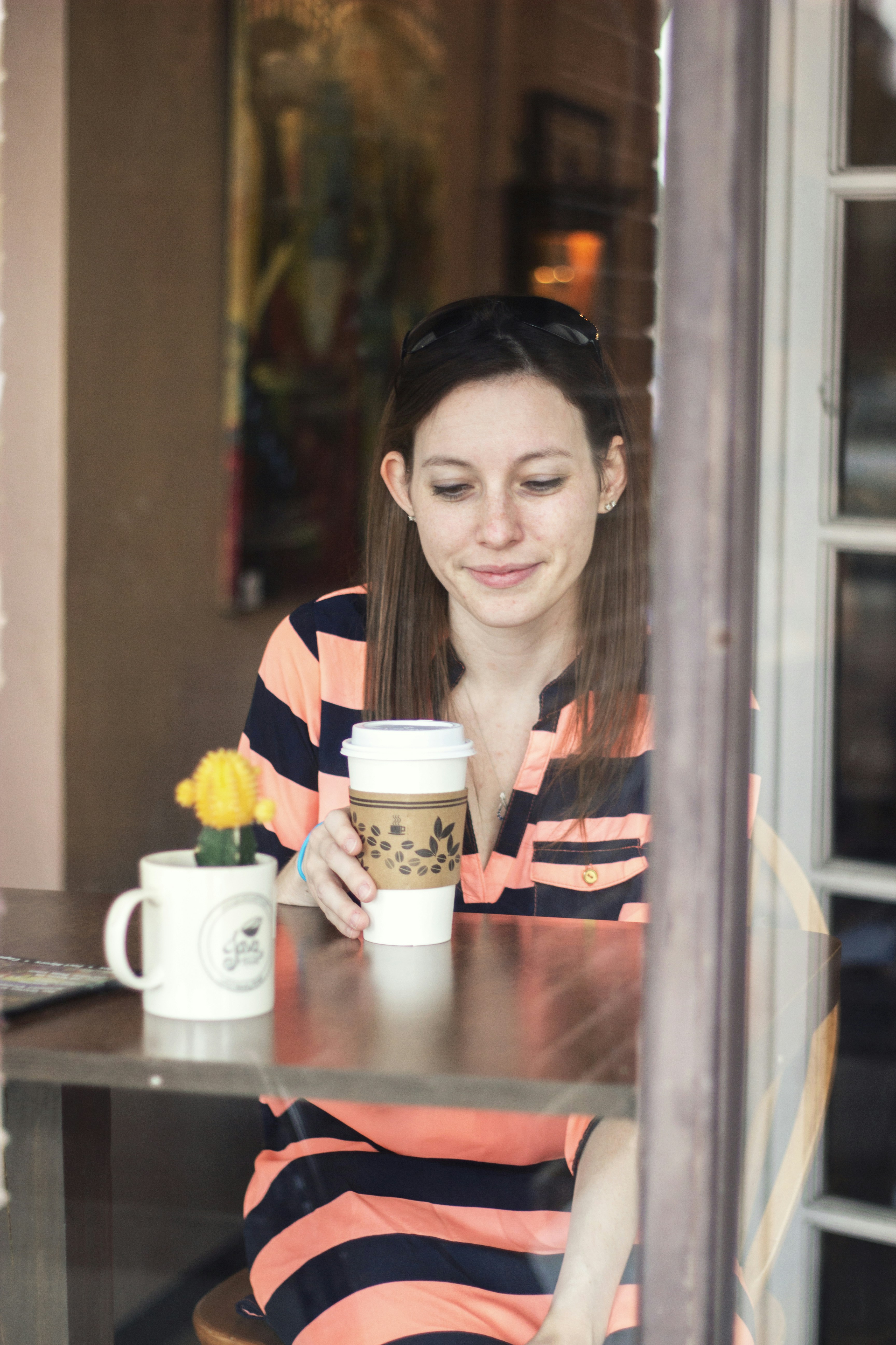 Woman in a striped dress enjoying a coffee at a café table, with a small potted plant and a magazine nearby.