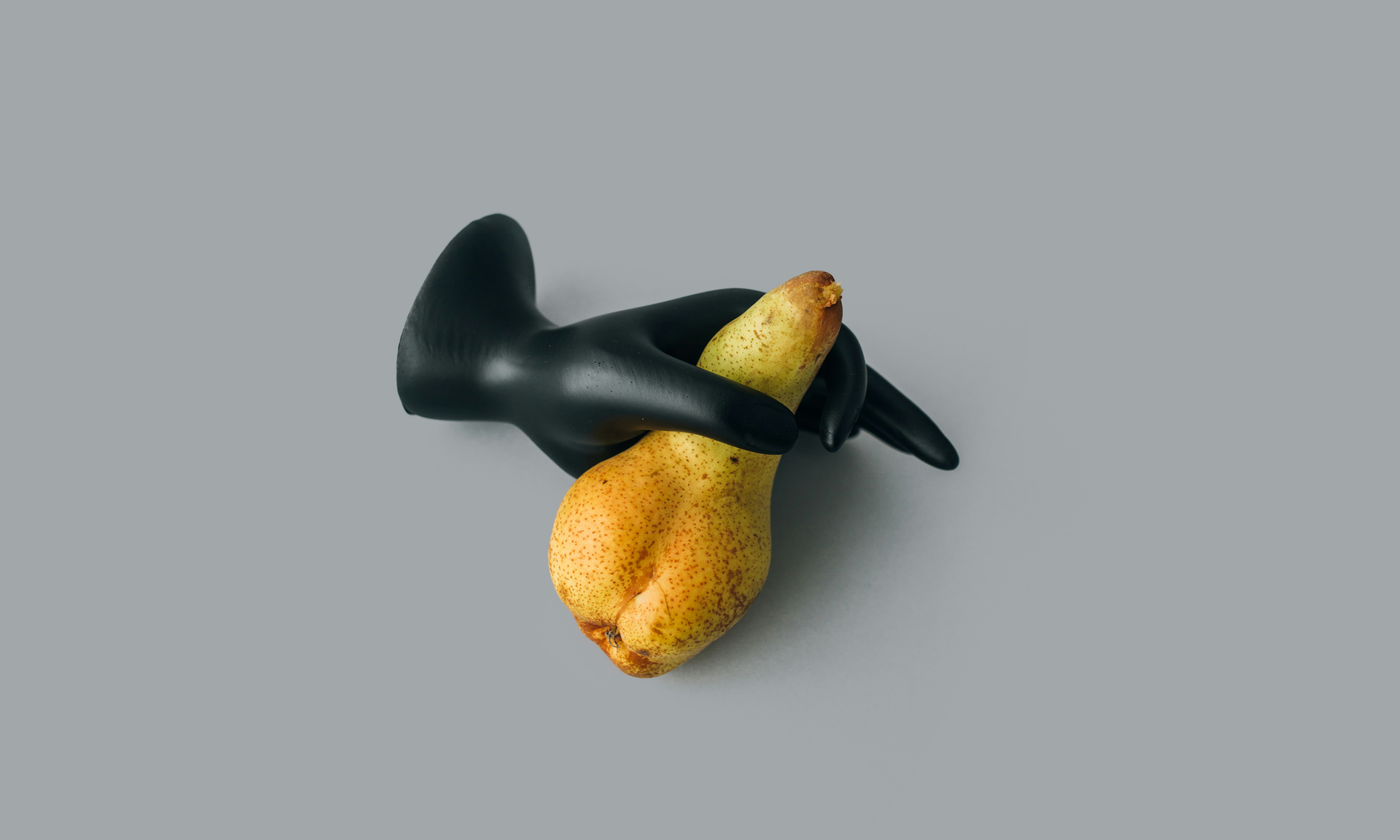 A black-gloved hand gently cradles a ripe pear against a muted gray background, highlighting the interplay of textures and colors.