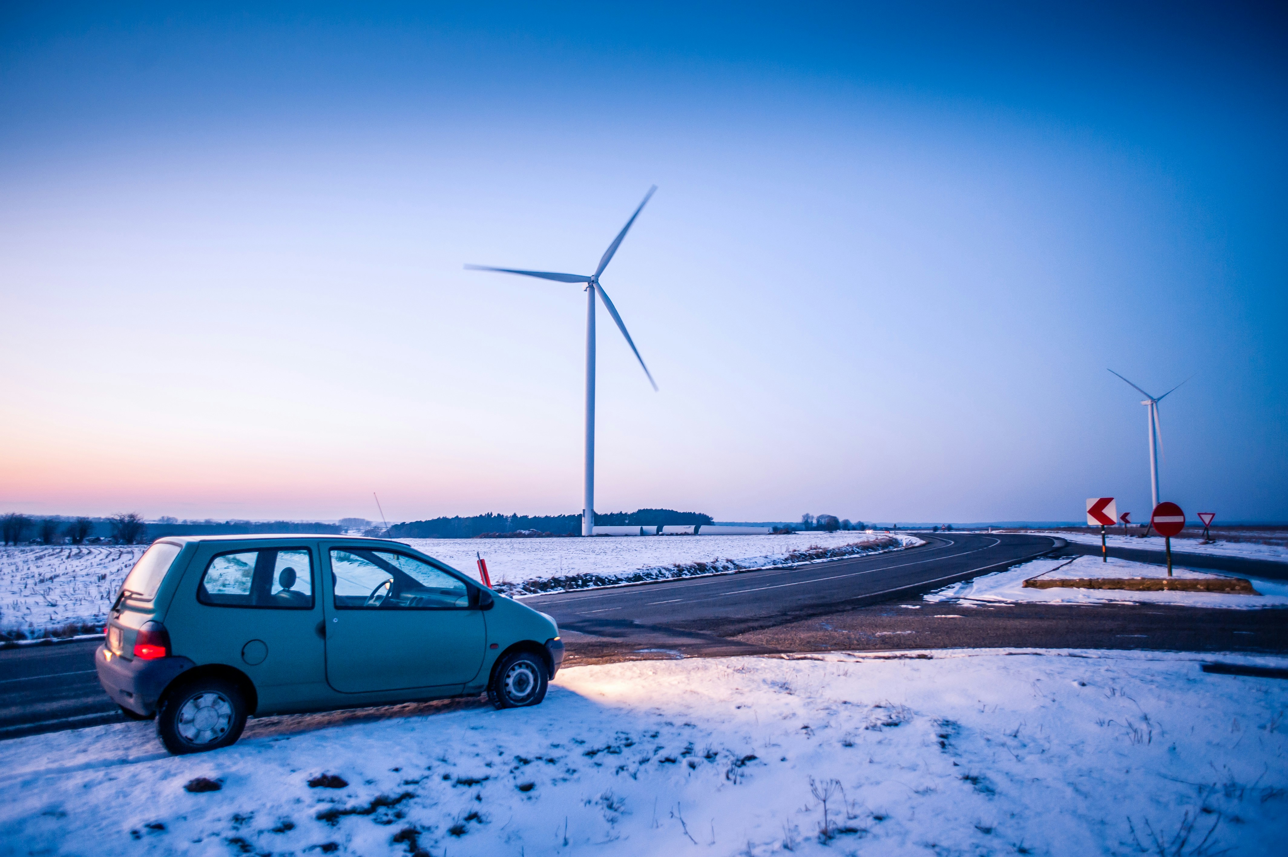 Crossroads in Cold Light: Blue Hatchback Beneath Wind Turbines