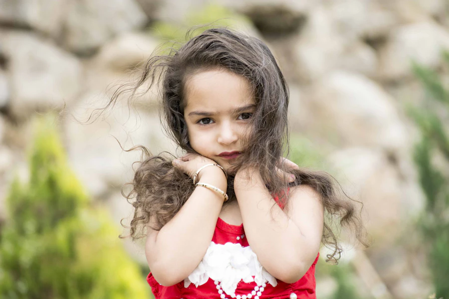 Young girl with flowing hair and a red dress poses thoughtfully against a natural backdrop. Her expression conveys a mix of curiosity and contemplation.