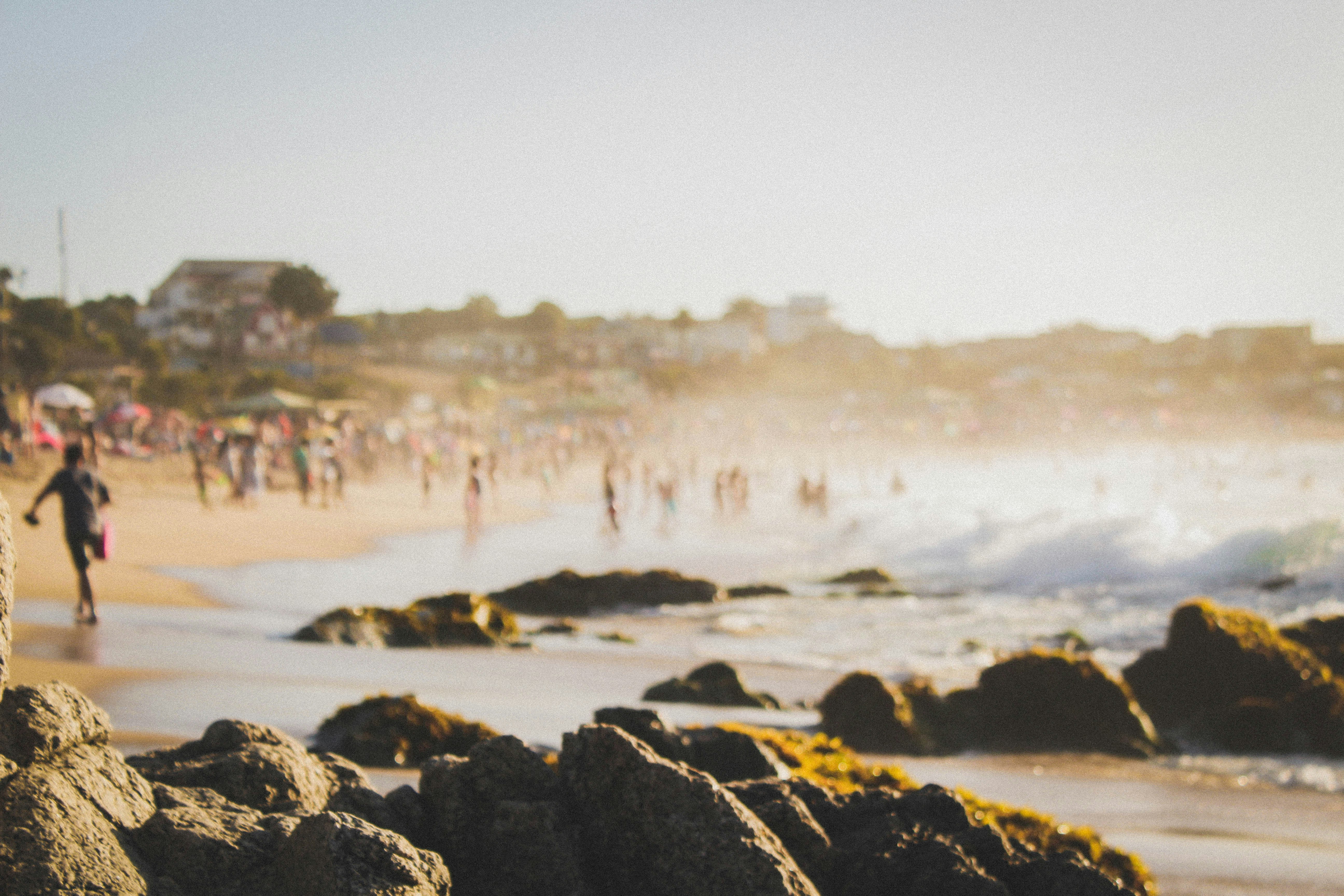 Crowded beach scene with people enjoying the sun and waves, framed by rocky foreground and distant seaside homes.