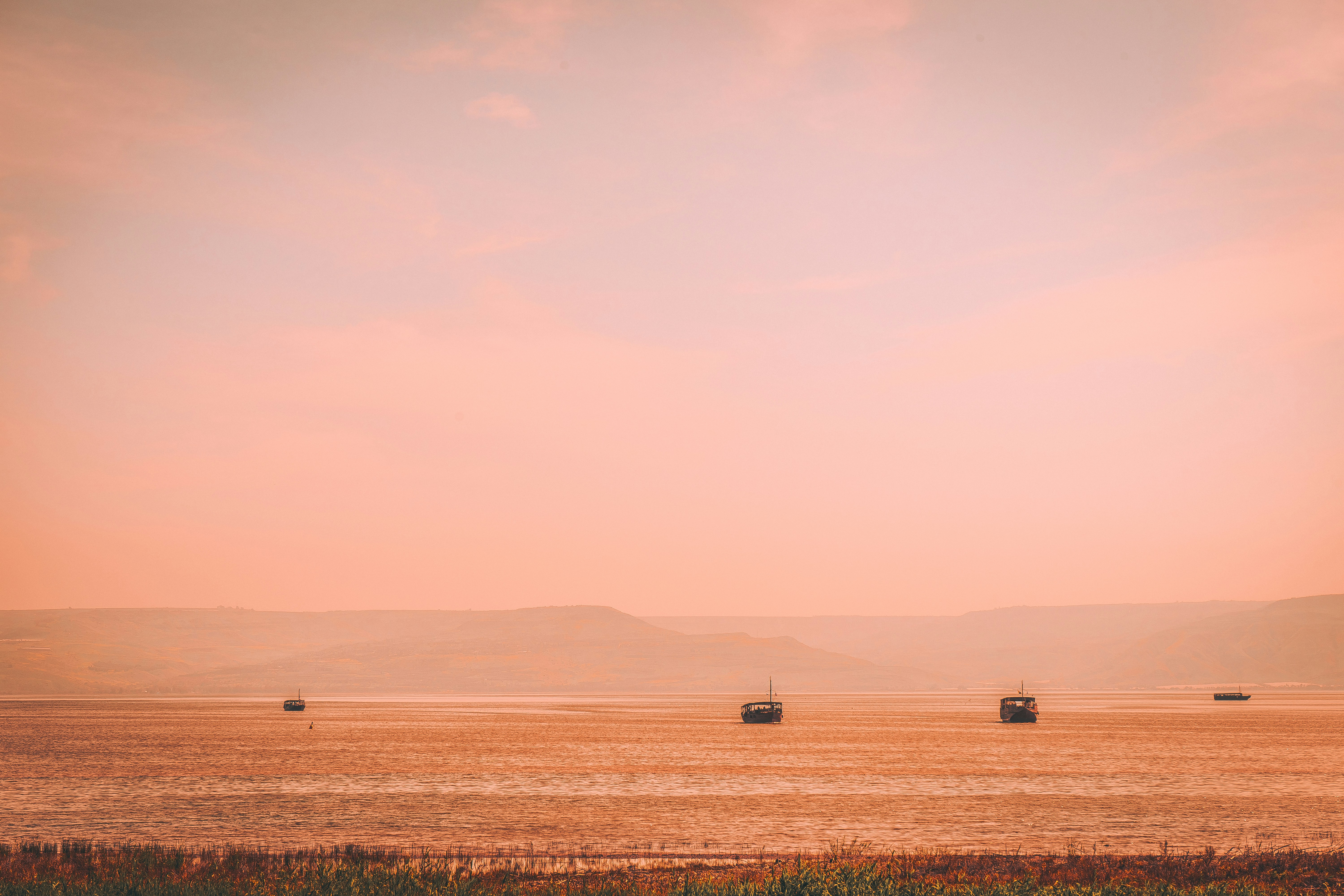 Boats drift on the calm Sea of Galilee under a soft, pastel sky.