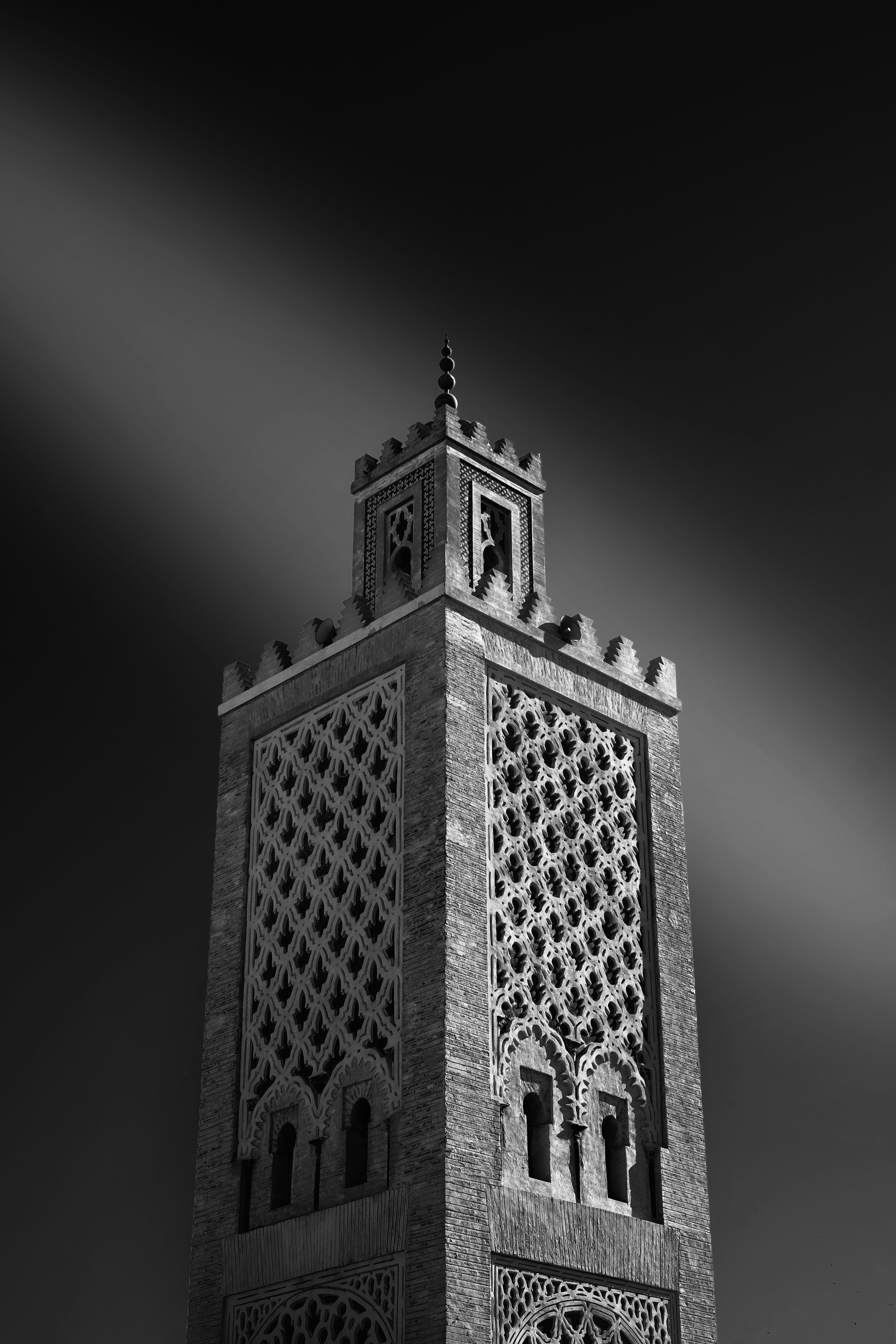 Intricate minaret adorned with geometric patterns rises against a dramatic sky, showcasing architectural elegance in monochrome.